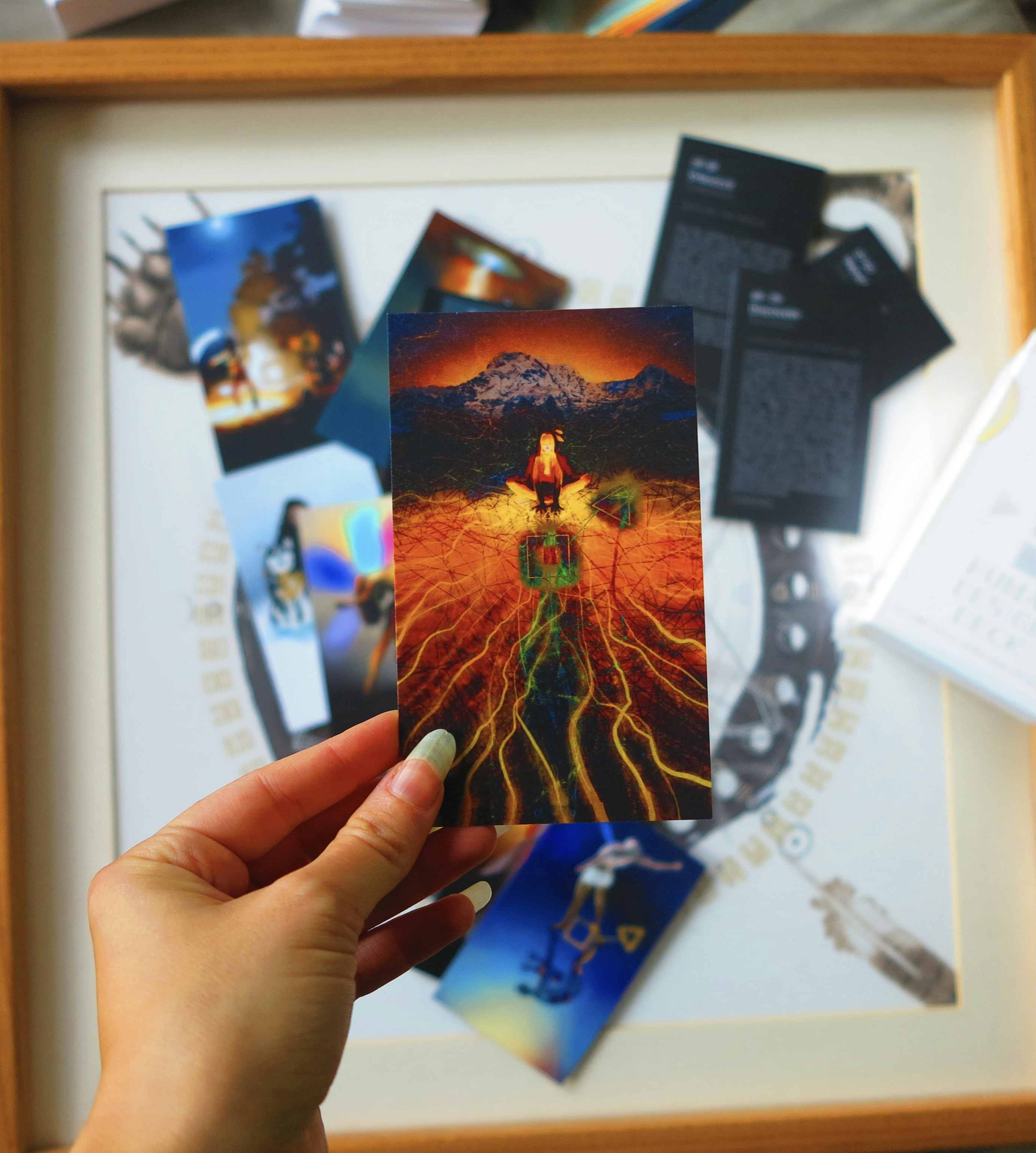 A hand holds a vibrant postcard featuring a surreal landscape with a figure standing amidst flowing energy lines, surrounded by various cards and a decorative frame.