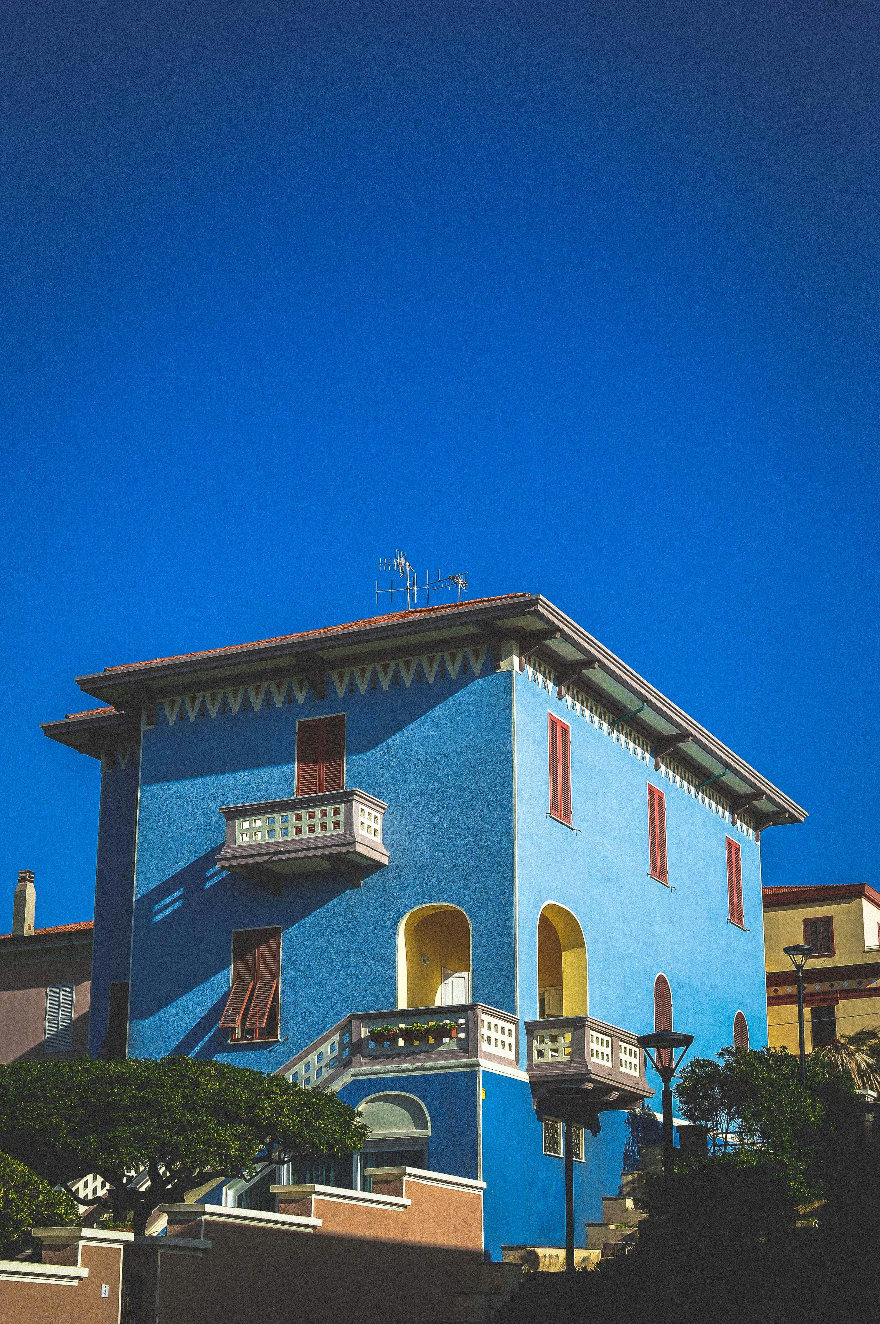 Colorful blue house with distinctive architectural features and balconies, surrounded by greenery and a clear blue sky.