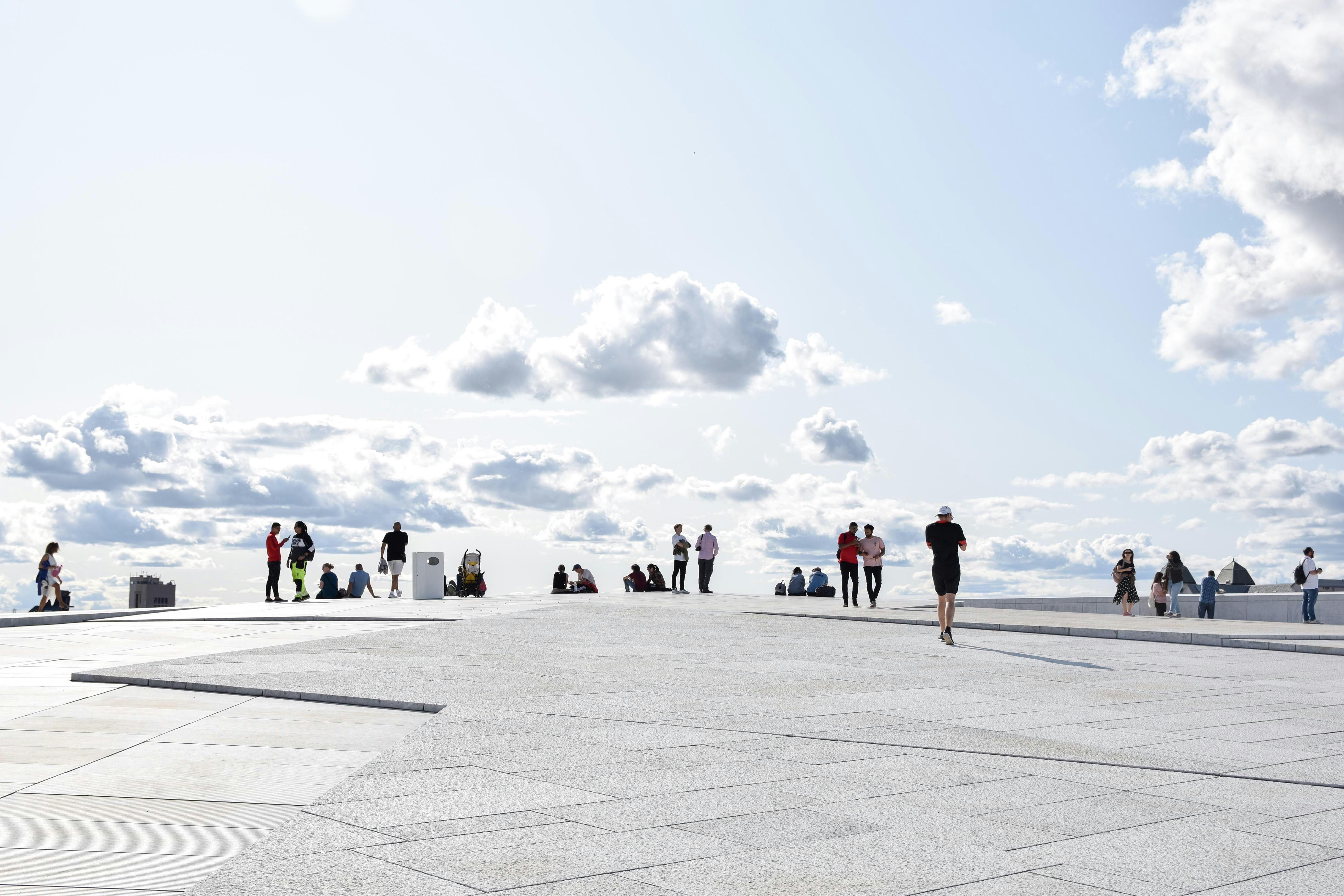 Silhouetted figures walking across a vast concrete expanse under a bright sky with scattered clouds.