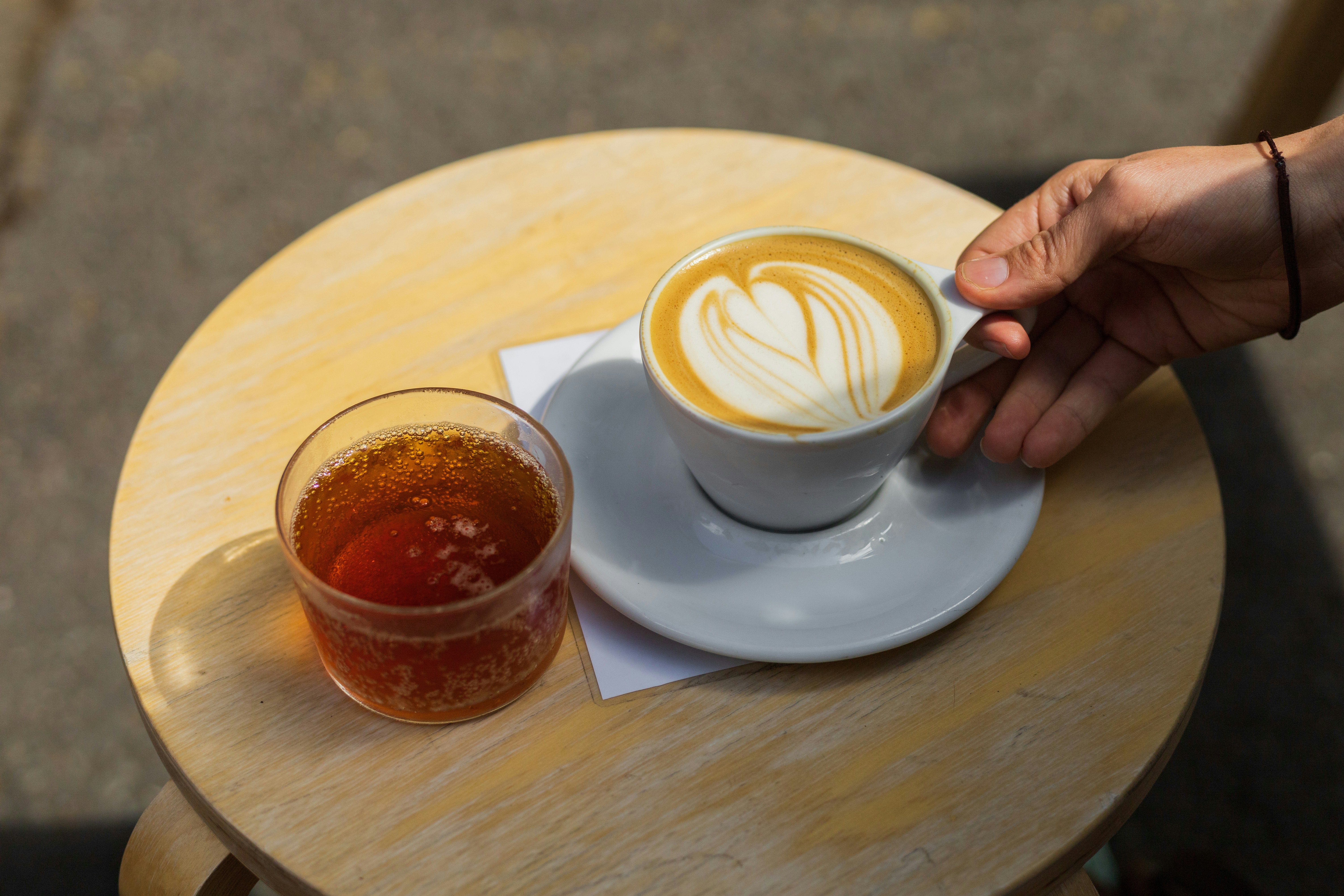 Latte Heart on a Sunlit Café Table, Framed by Hand and a Glass