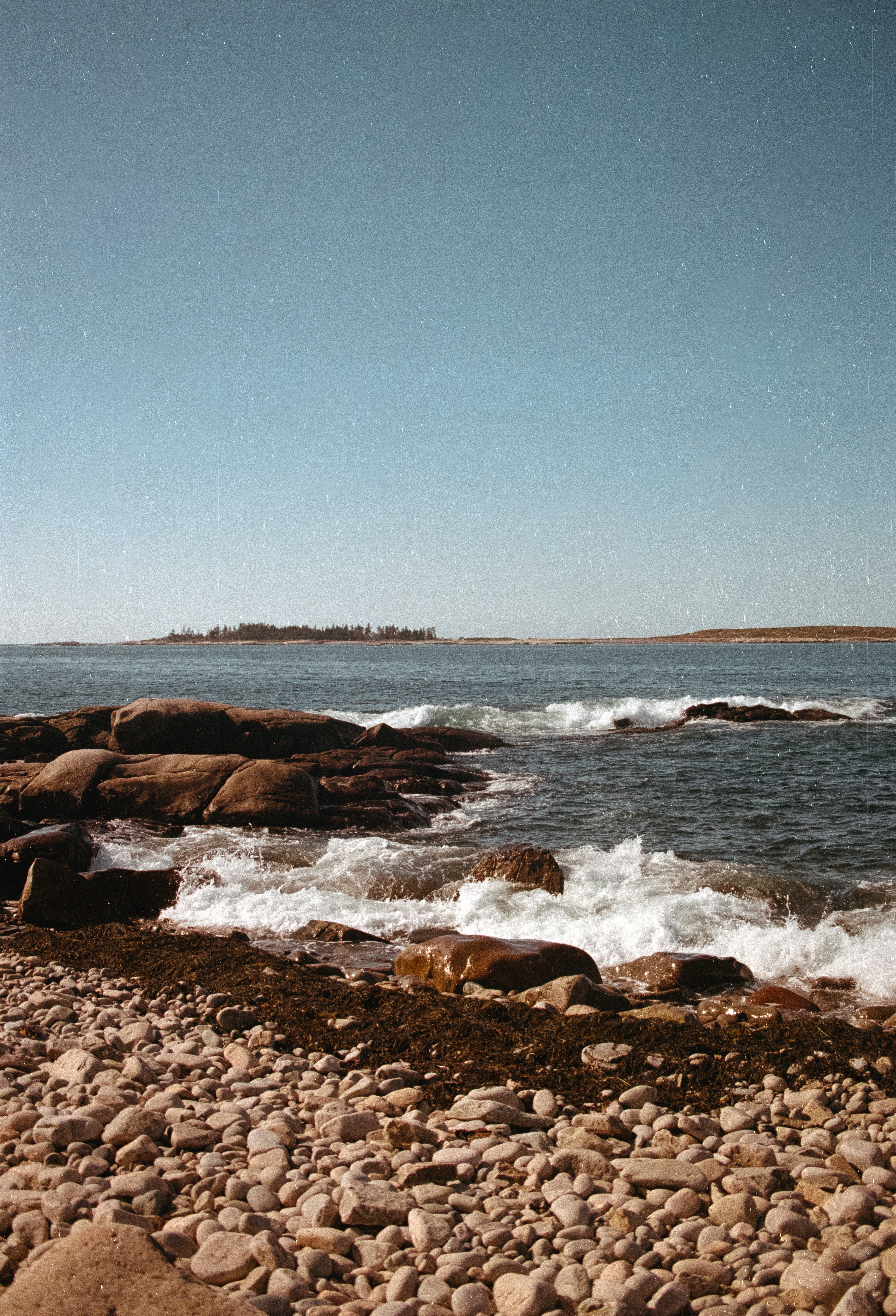 Waves gently lap against textured rocks along a pebbled shoreline, with a distant island silhouetted against a clear sky.