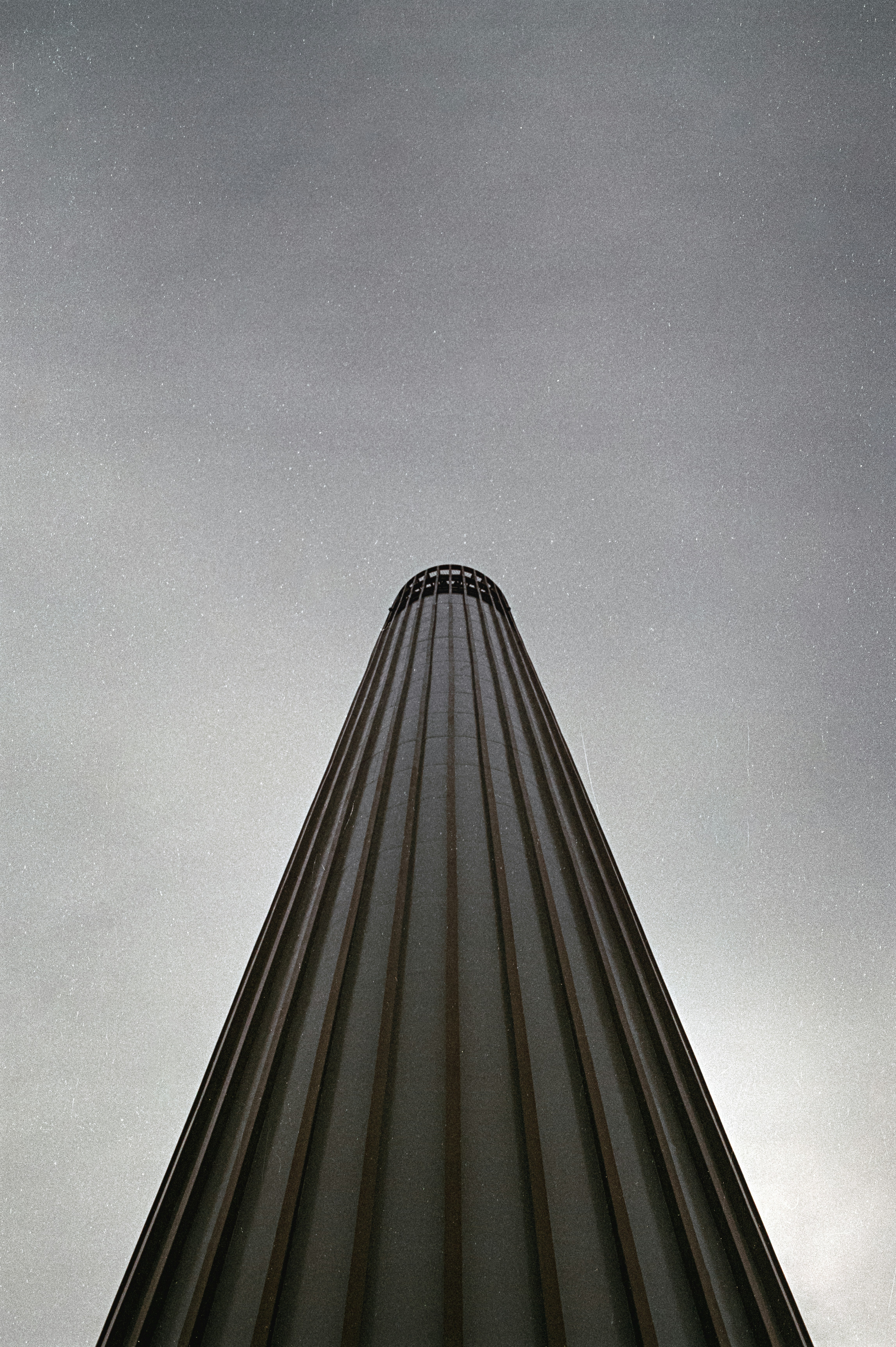 A tall, cylindrical structure reaching towards a cloudy sky, showcasing its textured surface and stark lines. The composition emphasizes height and architectural form.