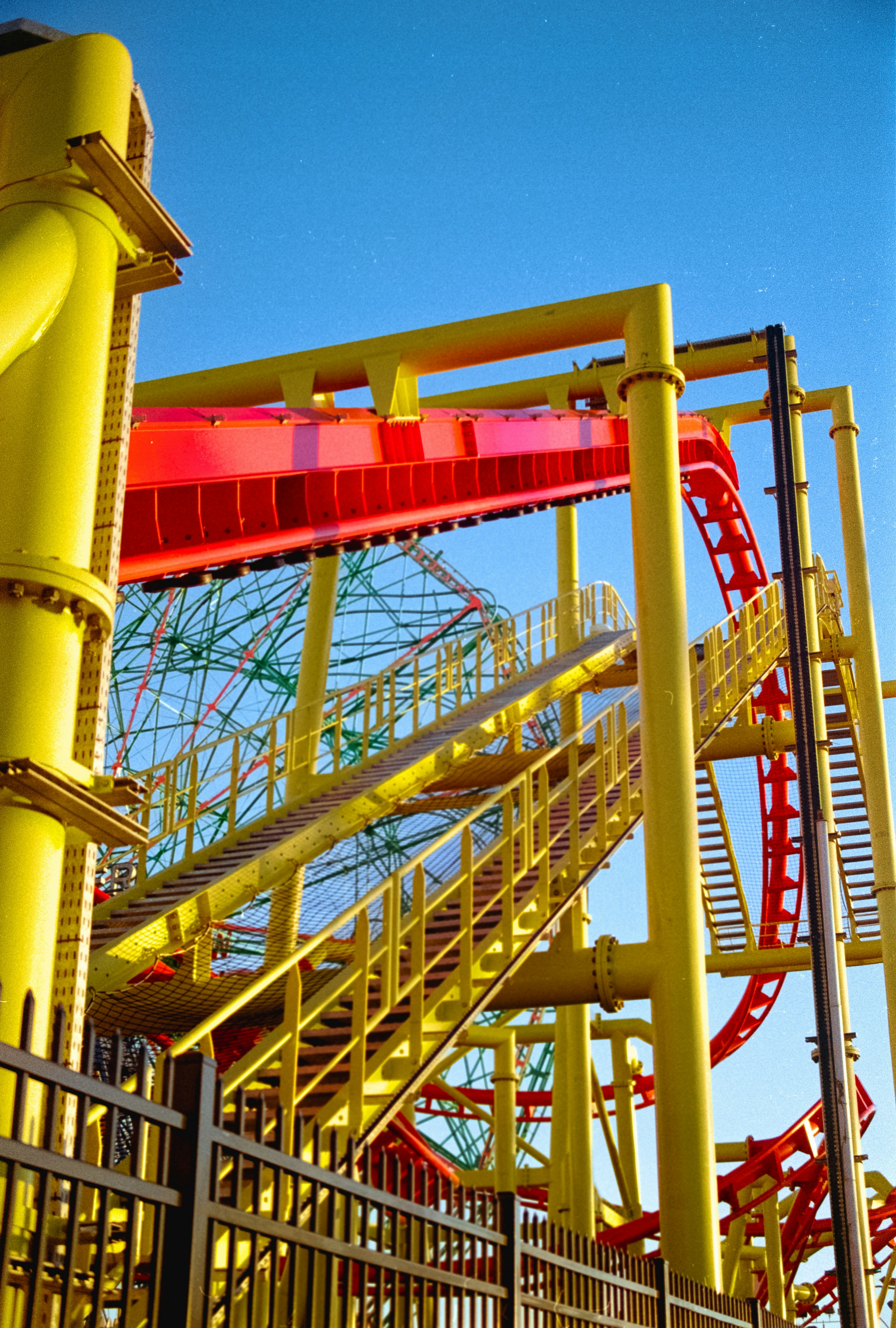 Colorful roller coaster structure with yellow and red tracks intertwined, showcasing the intricate design and engineering. The bright blue sky serves as a backdrop.