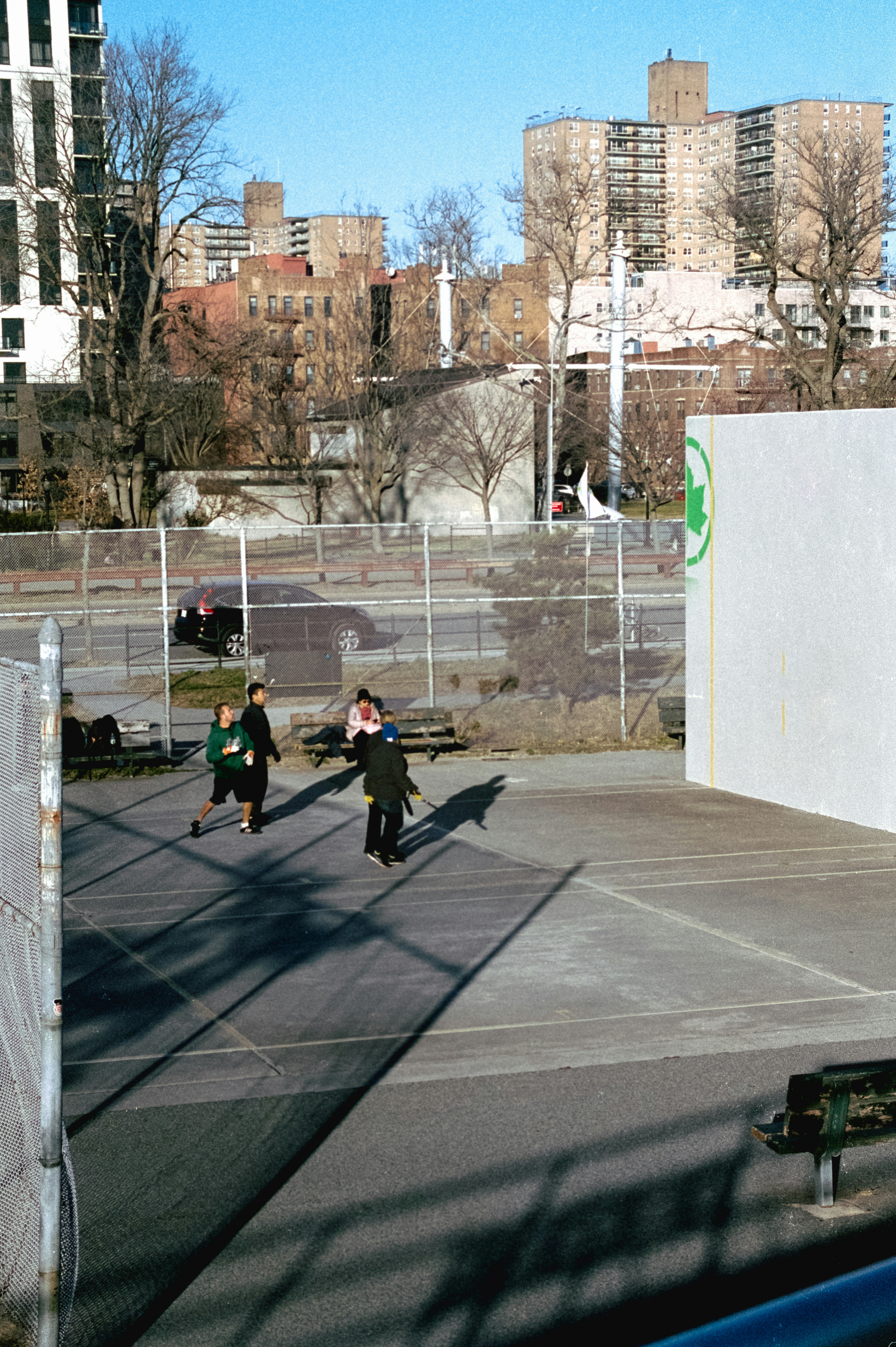 Children chase a ball on a sunlit concrete court, with a tall white wall to the right and an urban skyline in the distance.