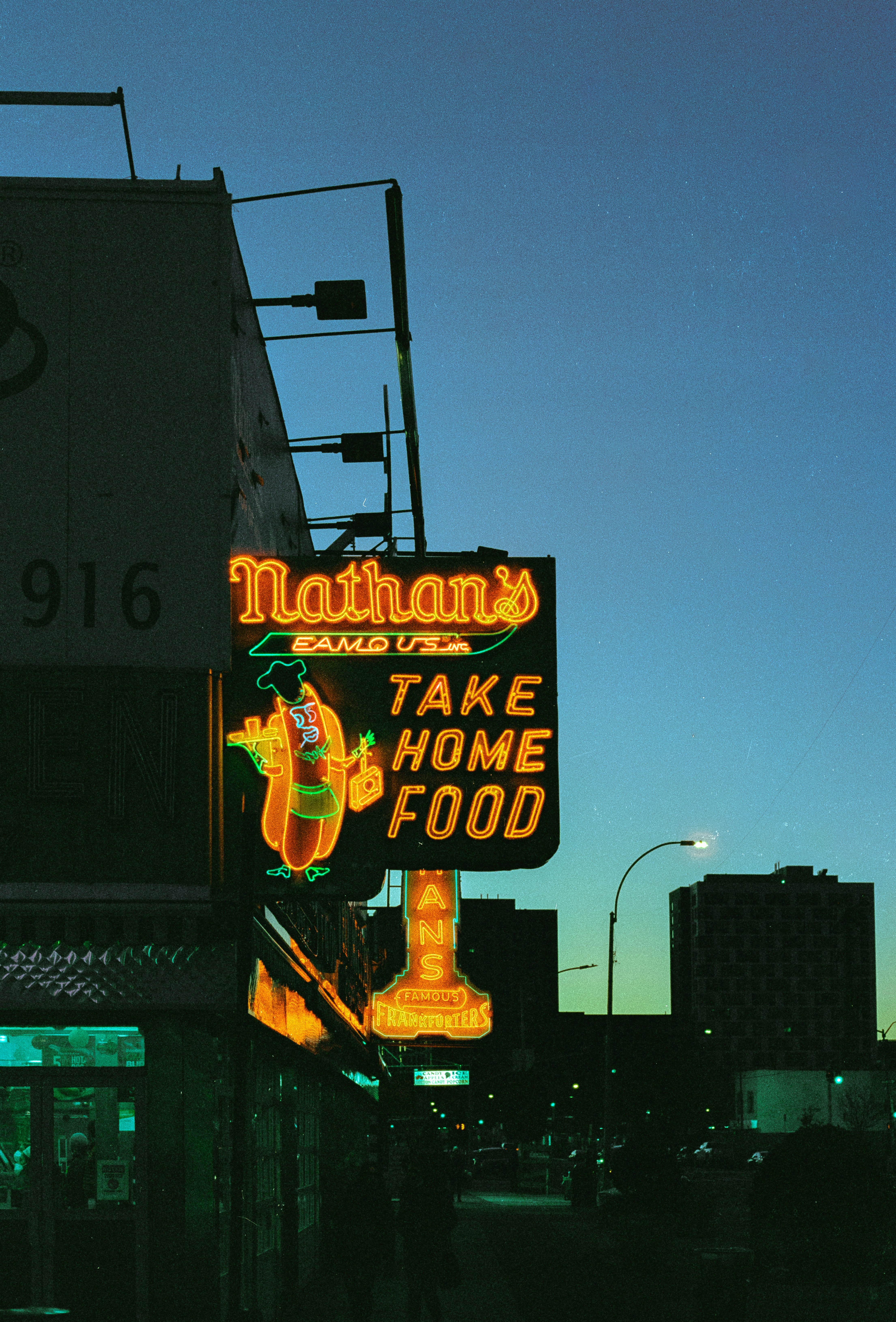 Neon logo for Nathan's Famous glows on a dusk-lit street, with a Take Home Food sign and silhouetted storefronts.