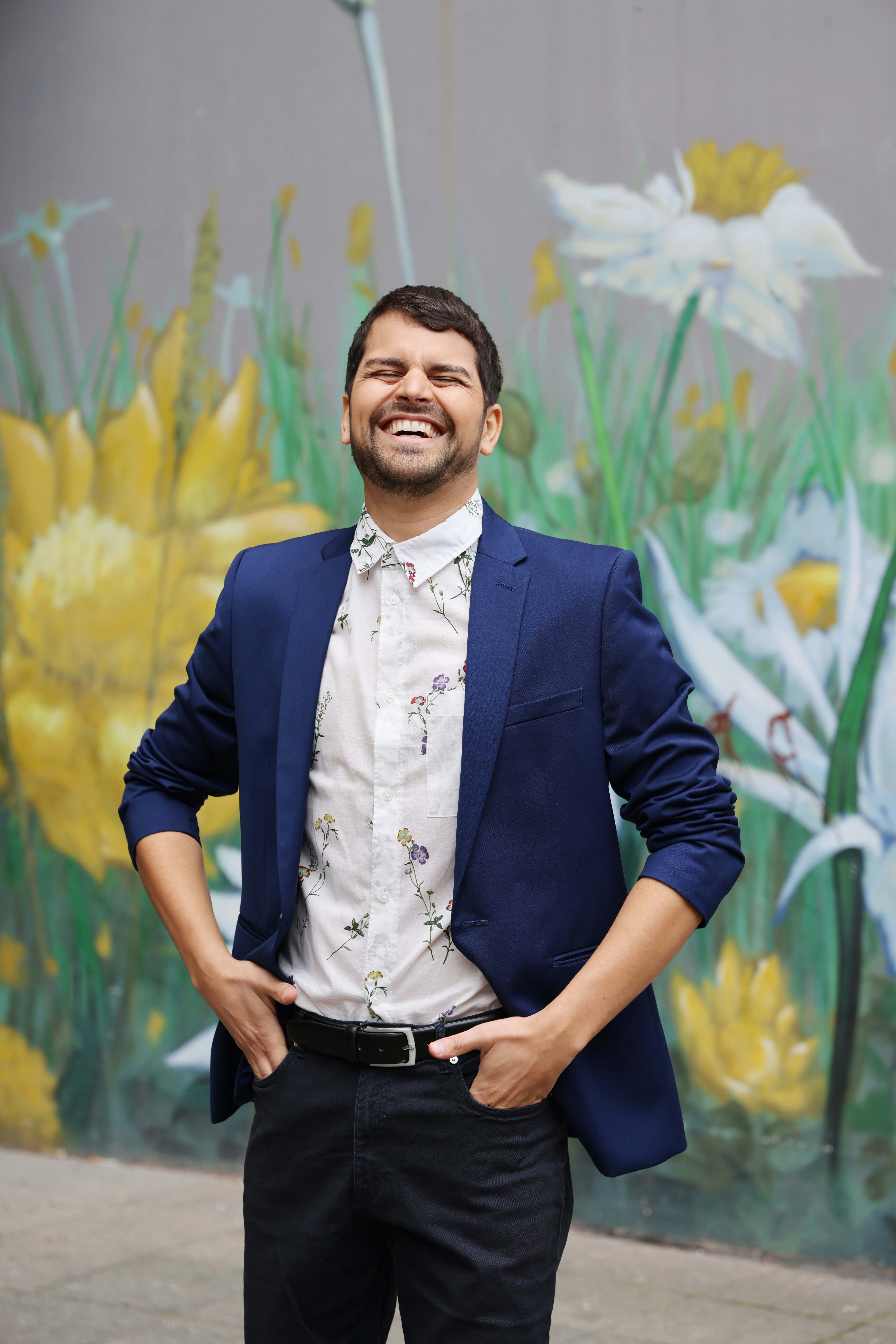 Man in a blue blazer and floral shirt smiles joyfully in front of a vibrant floral mural.