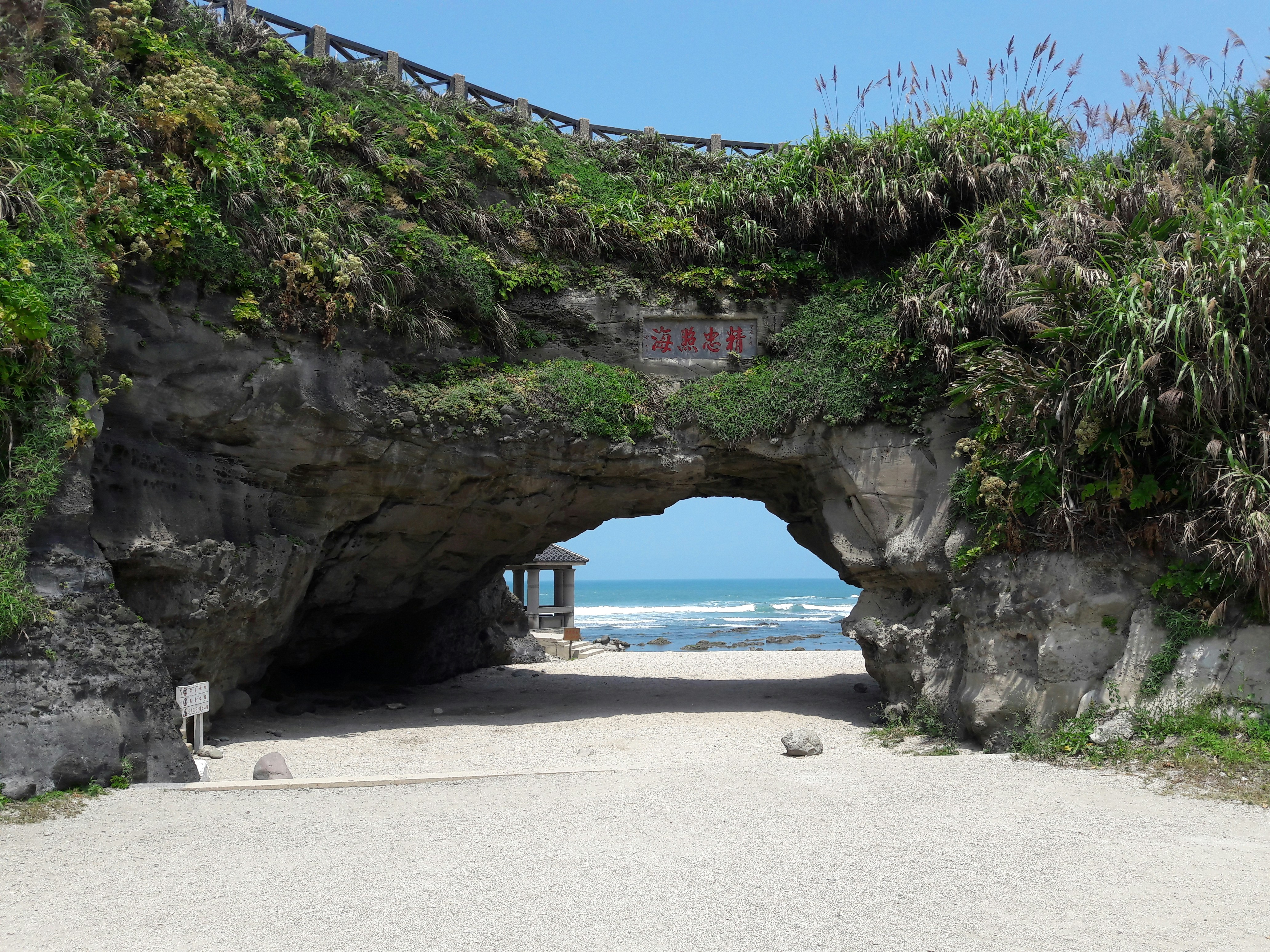 Natural rock archway framing a serene beach scene, with lush greenery and a distant lookout structure. The ocean waves gently lap at the shore.