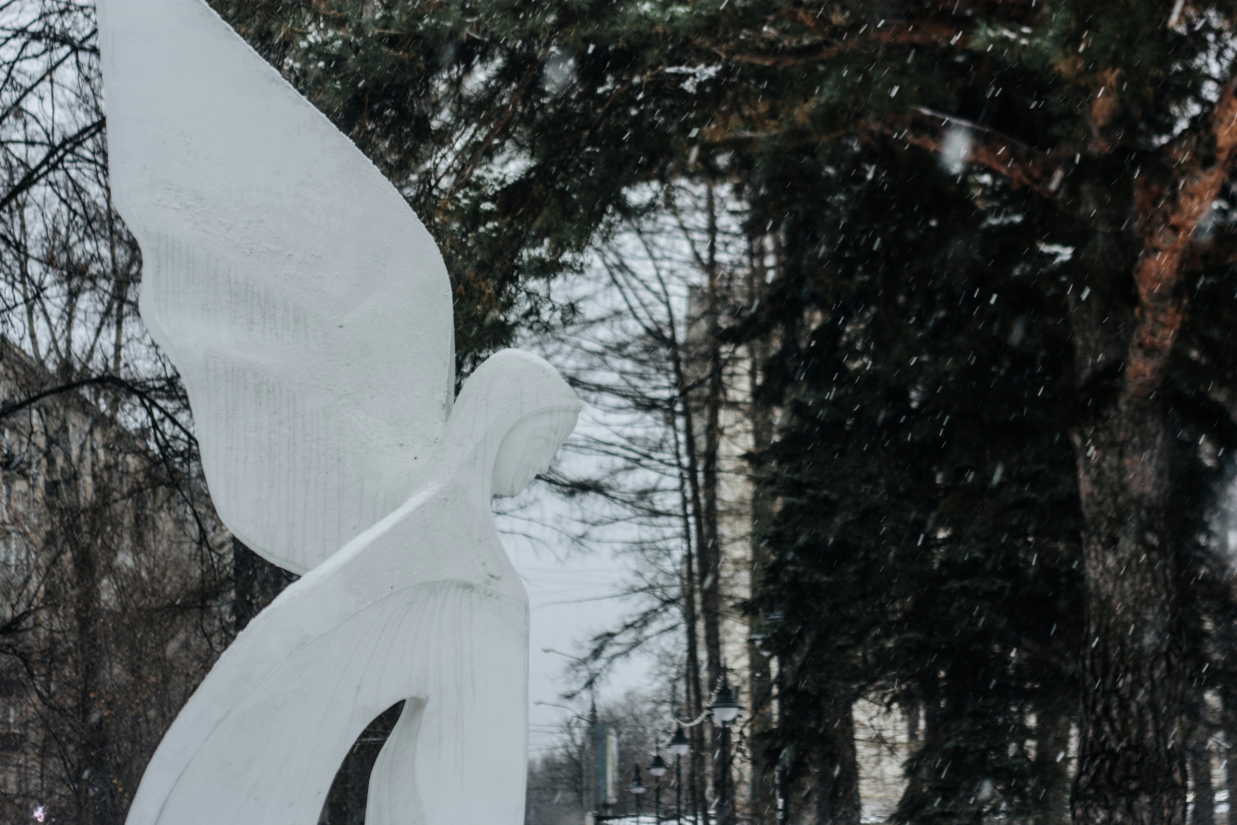 A white angel sculpture stands amidst falling snow, surrounded by tall trees in a winter landscape. The serene atmosphere evokes a sense of tranquility.
