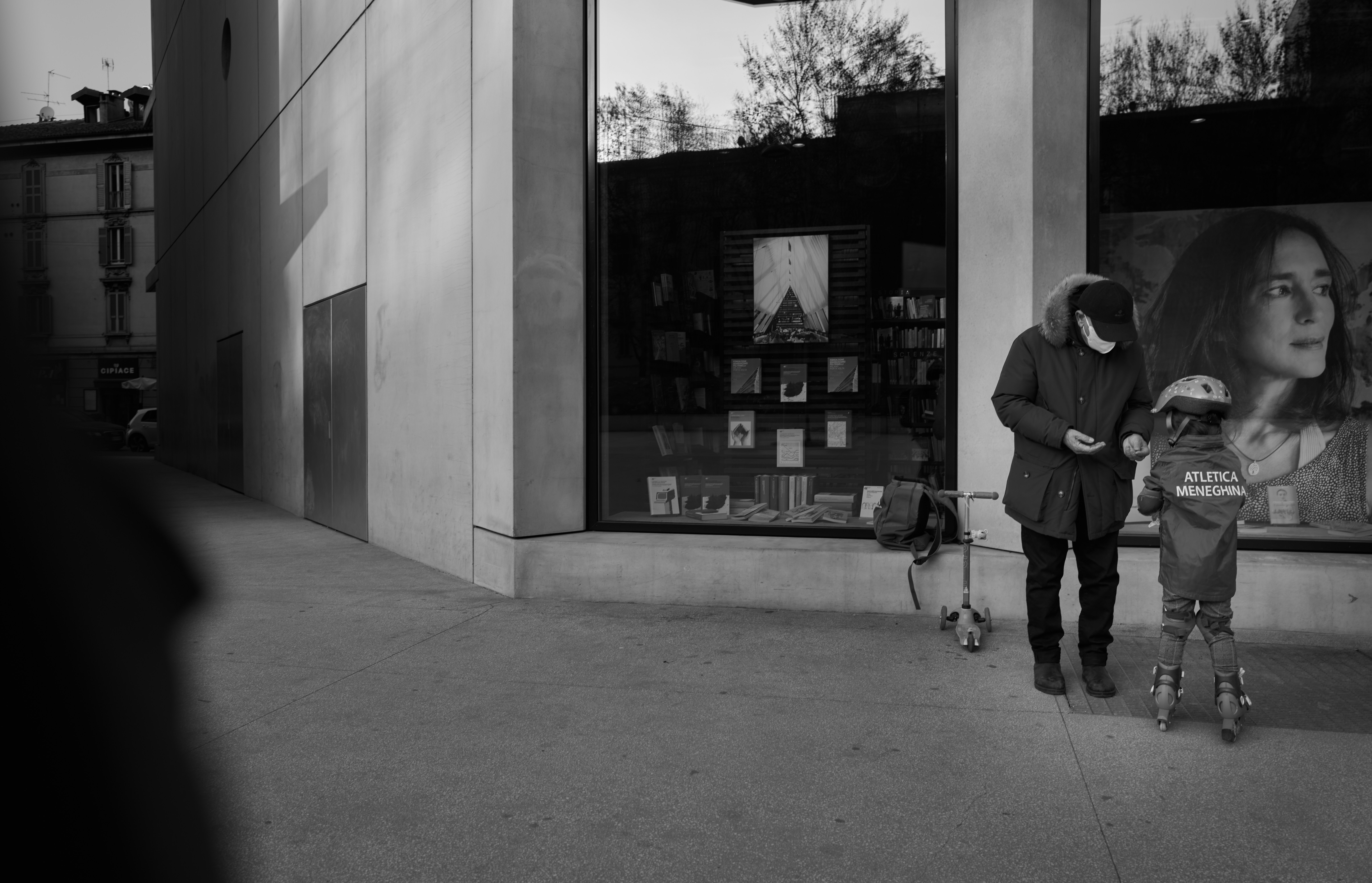 Monochrome street photograph of a man with a child beside a storefront window featuring a large portrait, with a small dog at their feet and a blurred silhouette in the foreground.