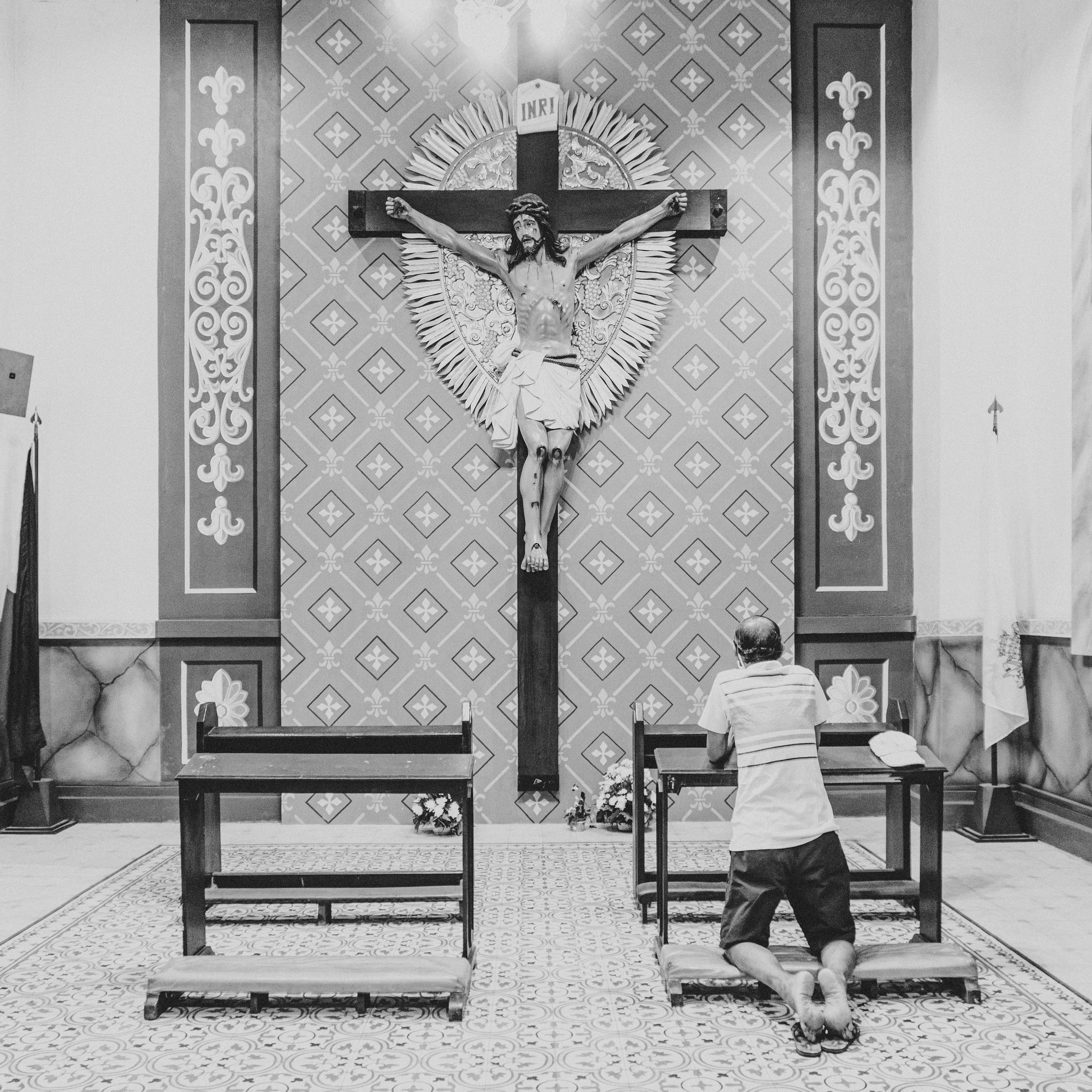 A man kneels in prayer before a crucifix adorned with angelic motifs in a serene chapel setting.