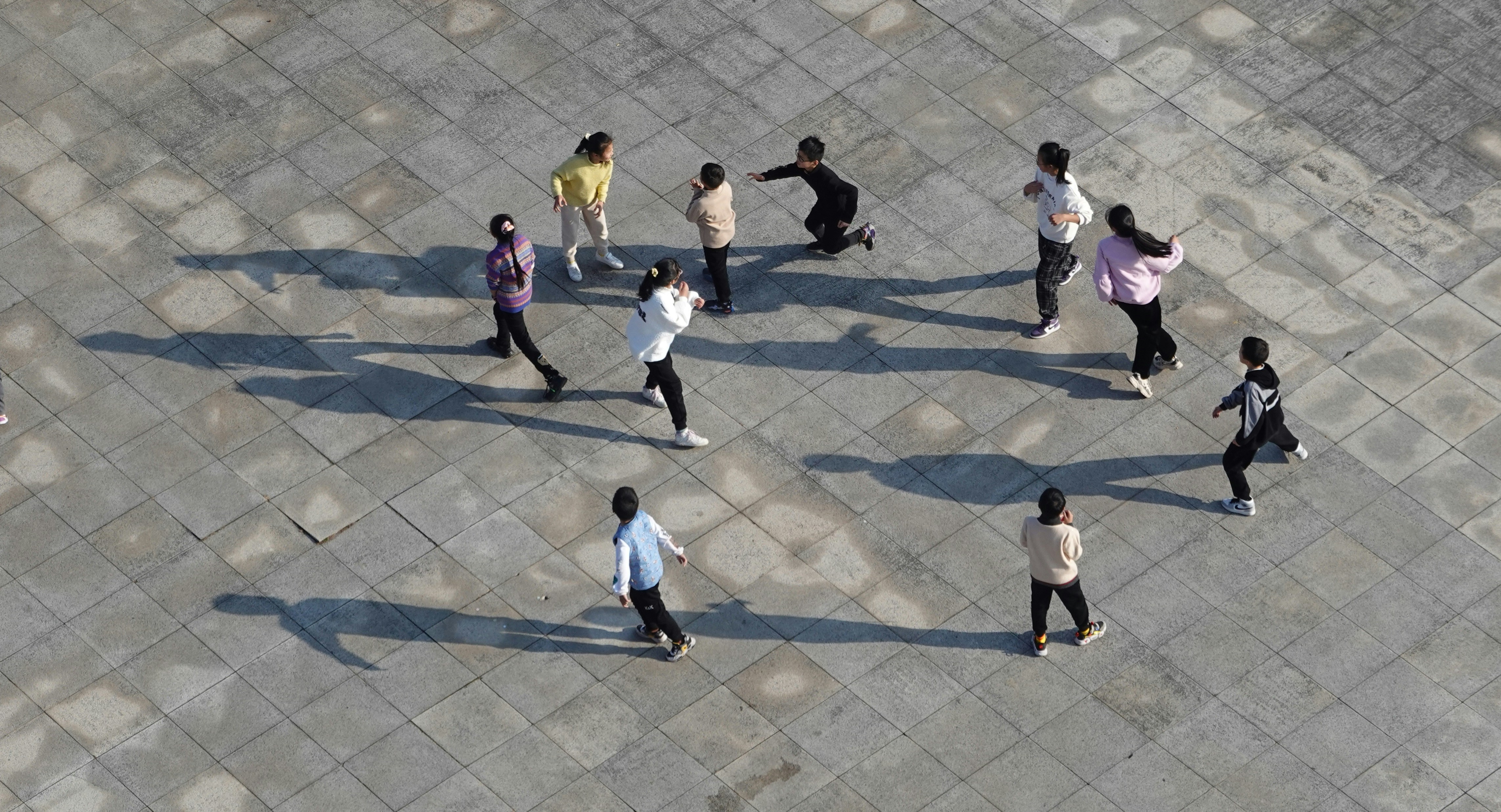 Group of individuals engaging in a synchronized dance on a sunlit plaza, casting elongated shadows on the ground.