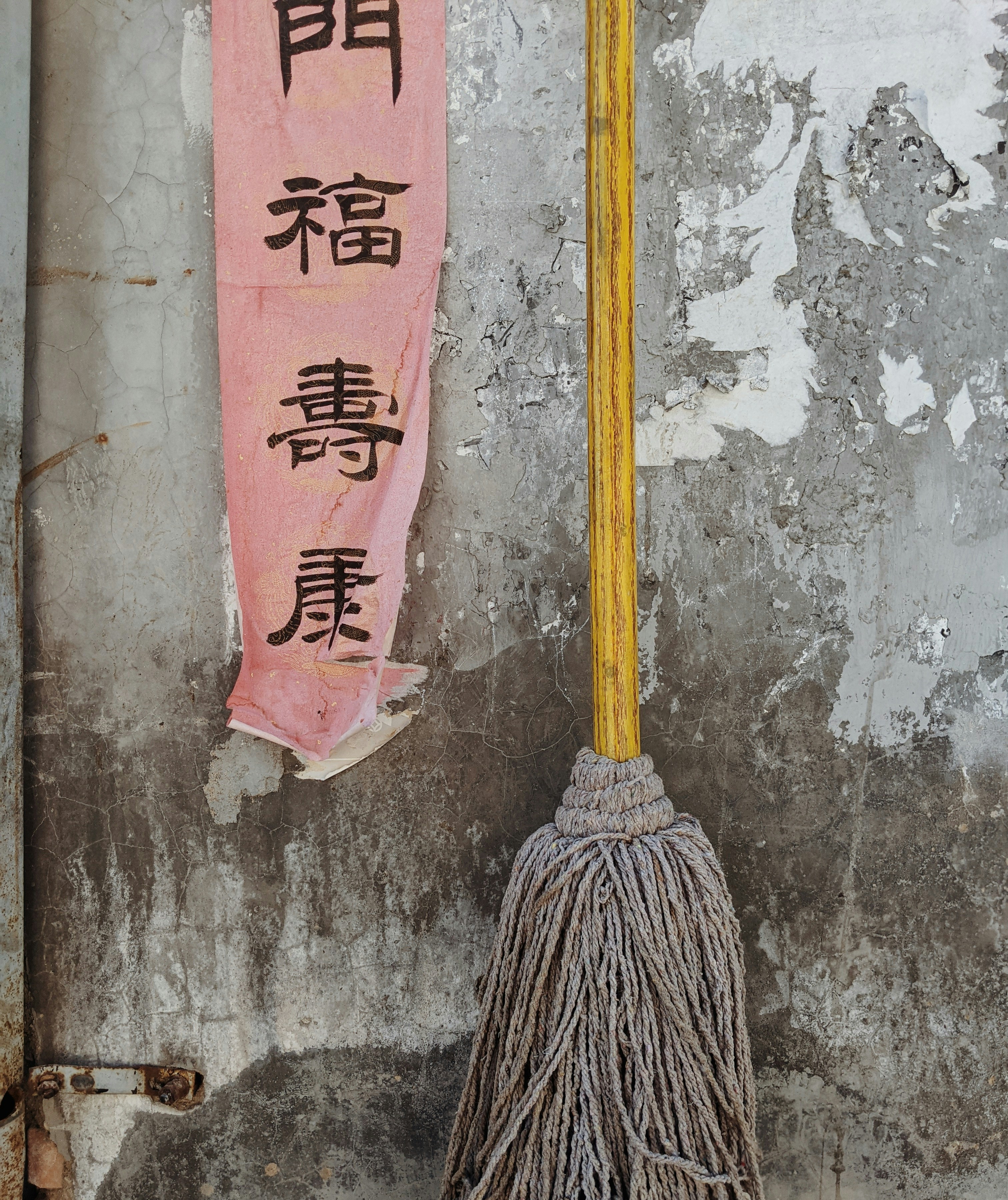 A weathered broom stands next to a faded pink banner with Chinese characters, set against a textured gray wall. The scene evokes a sense of history and cultural significance.