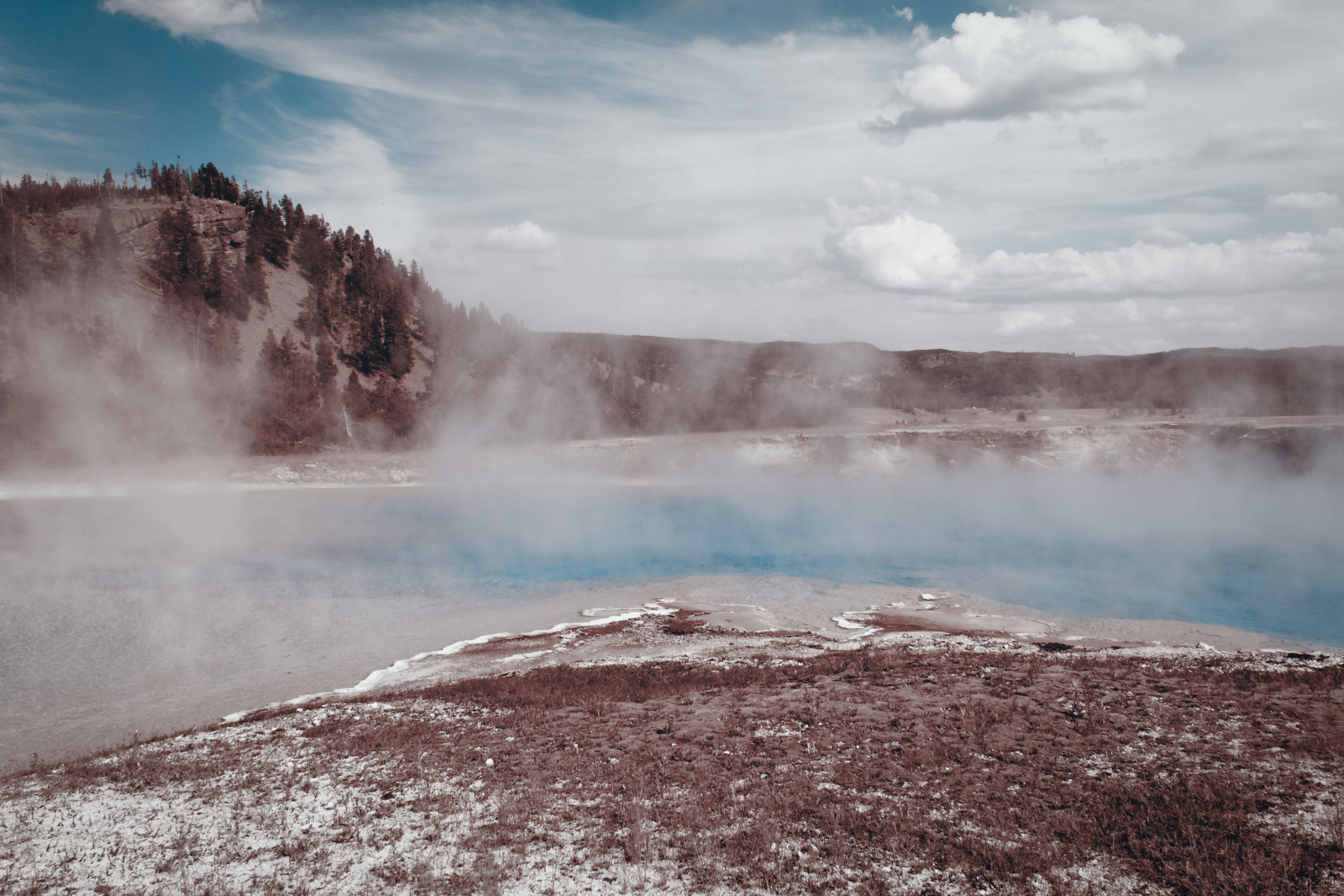 Steam rises from a geothermal pool, surrounded by a rugged landscape and distant hills under a cloudy sky.