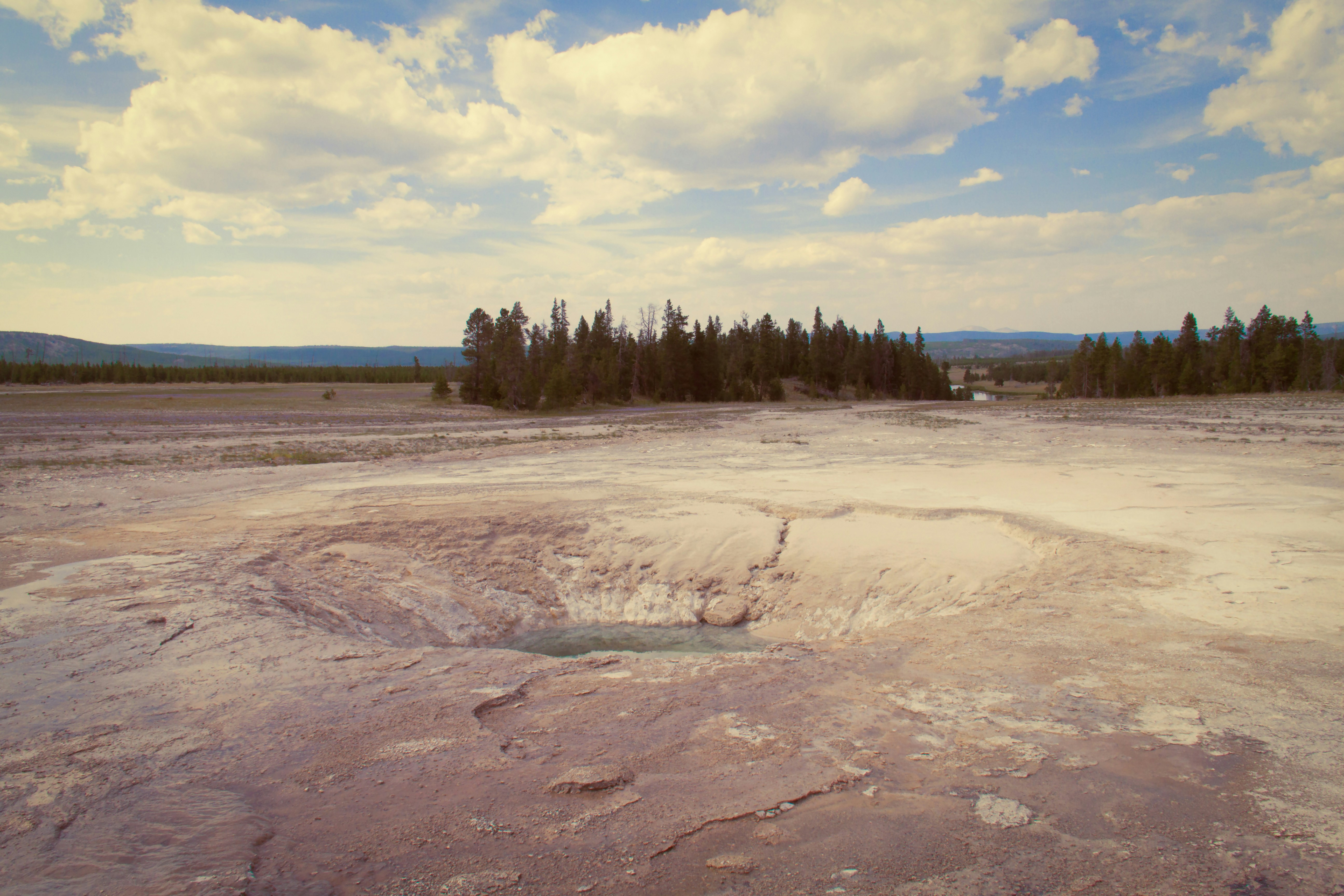 Geothermal feature with a circular depression revealing hints of turquoise water, surrounded by a rugged landscape and distant forested hills.