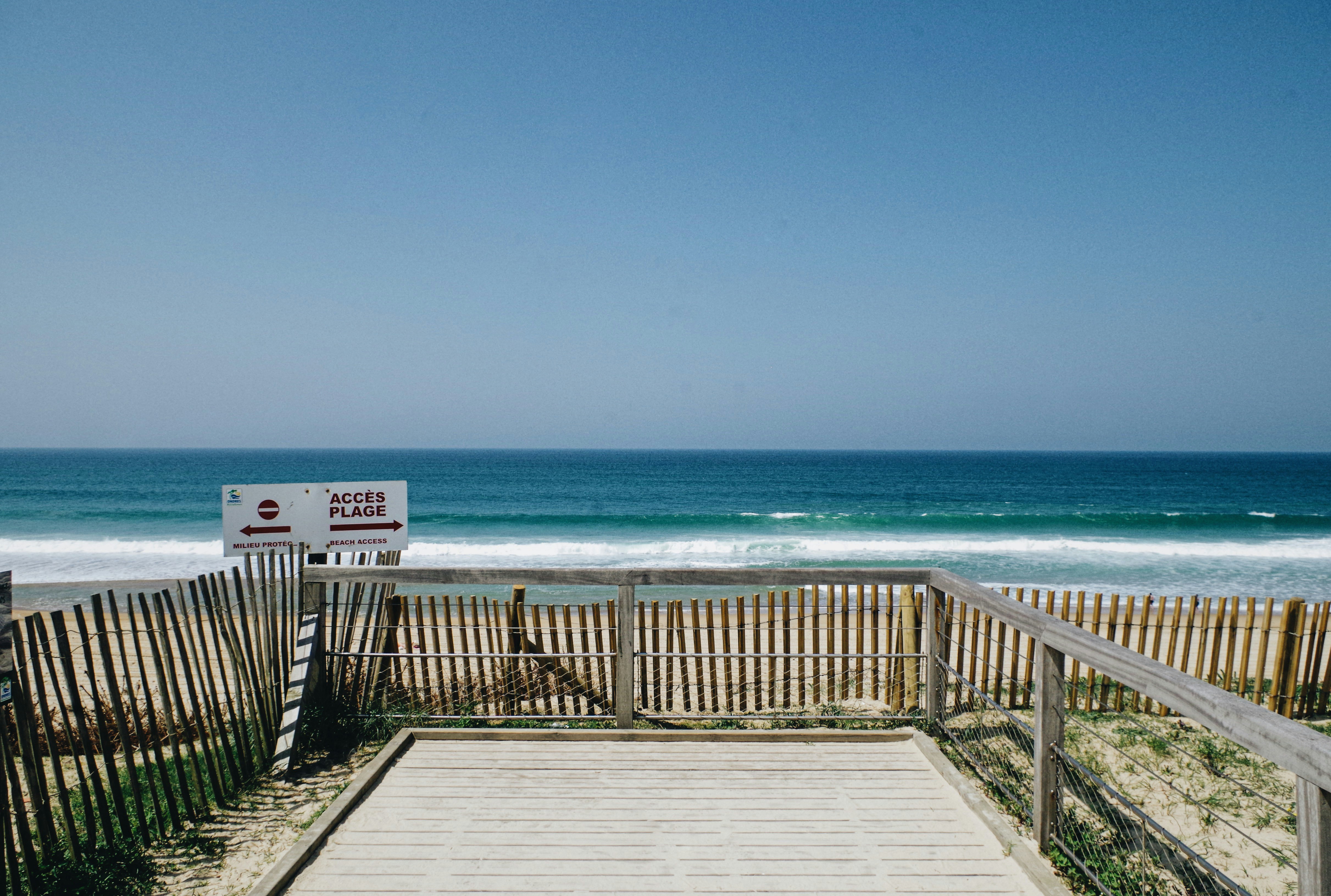Boardwalk leading to a calm beach and open ocean with a dune fence, sign, and clear sky in bright daylight.