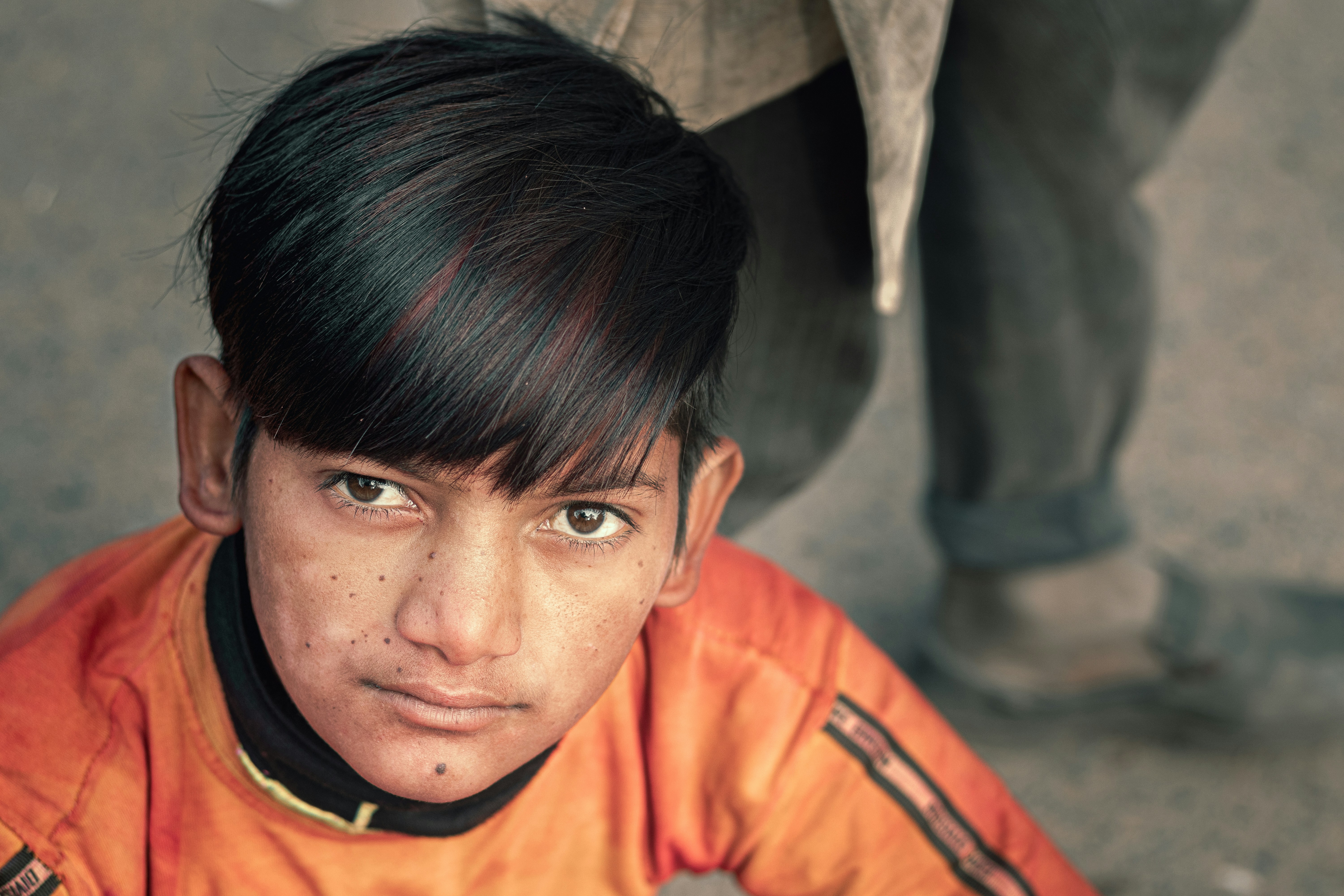 Young boy in an orange shirt gazing thoughtfully, surrounded by a blurred background, conveying a sense of resilience and curiosity.