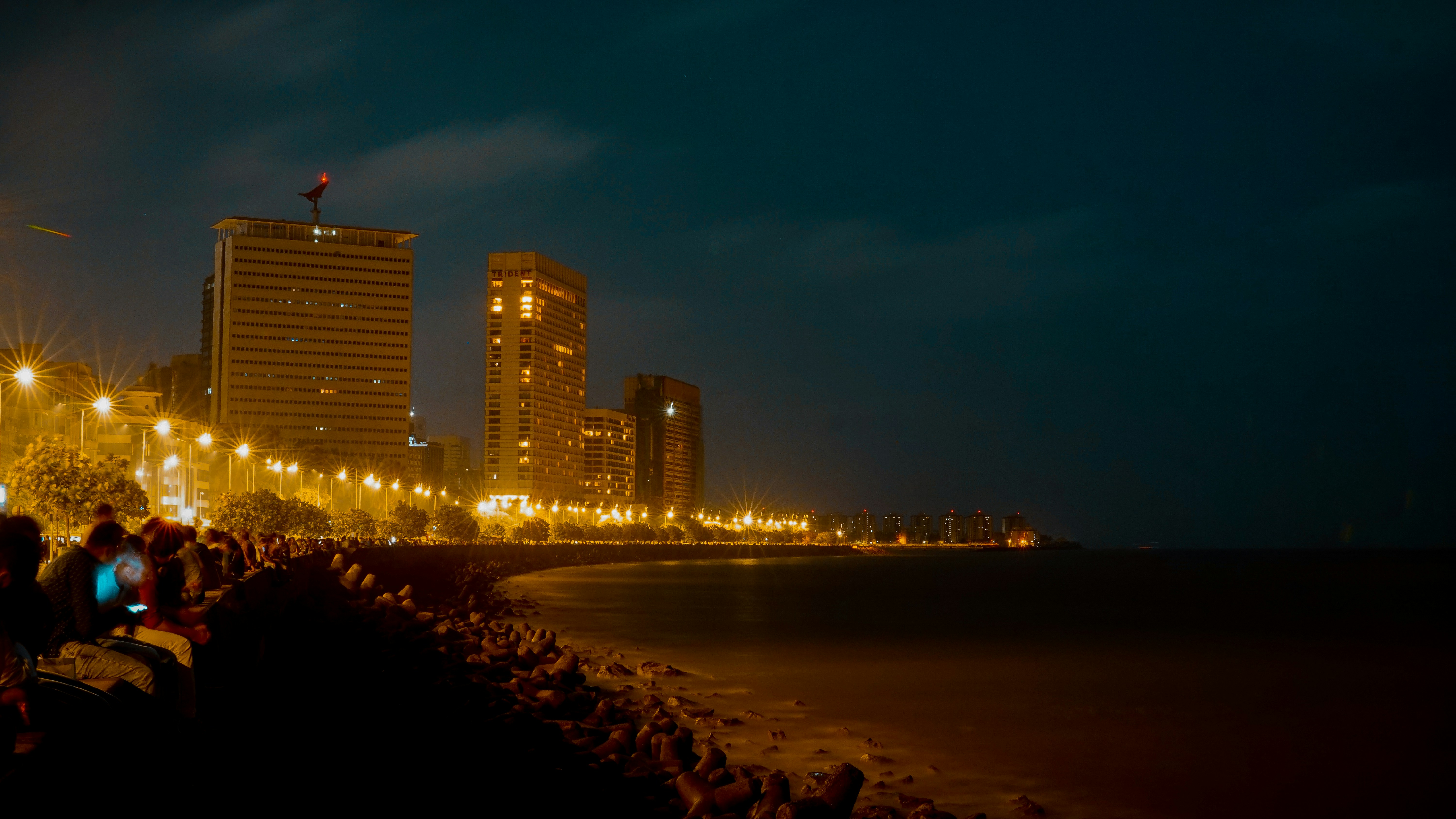 City skyline illuminated by streetlights along a coastal promenade at night, with silhouettes of people enjoying the view.