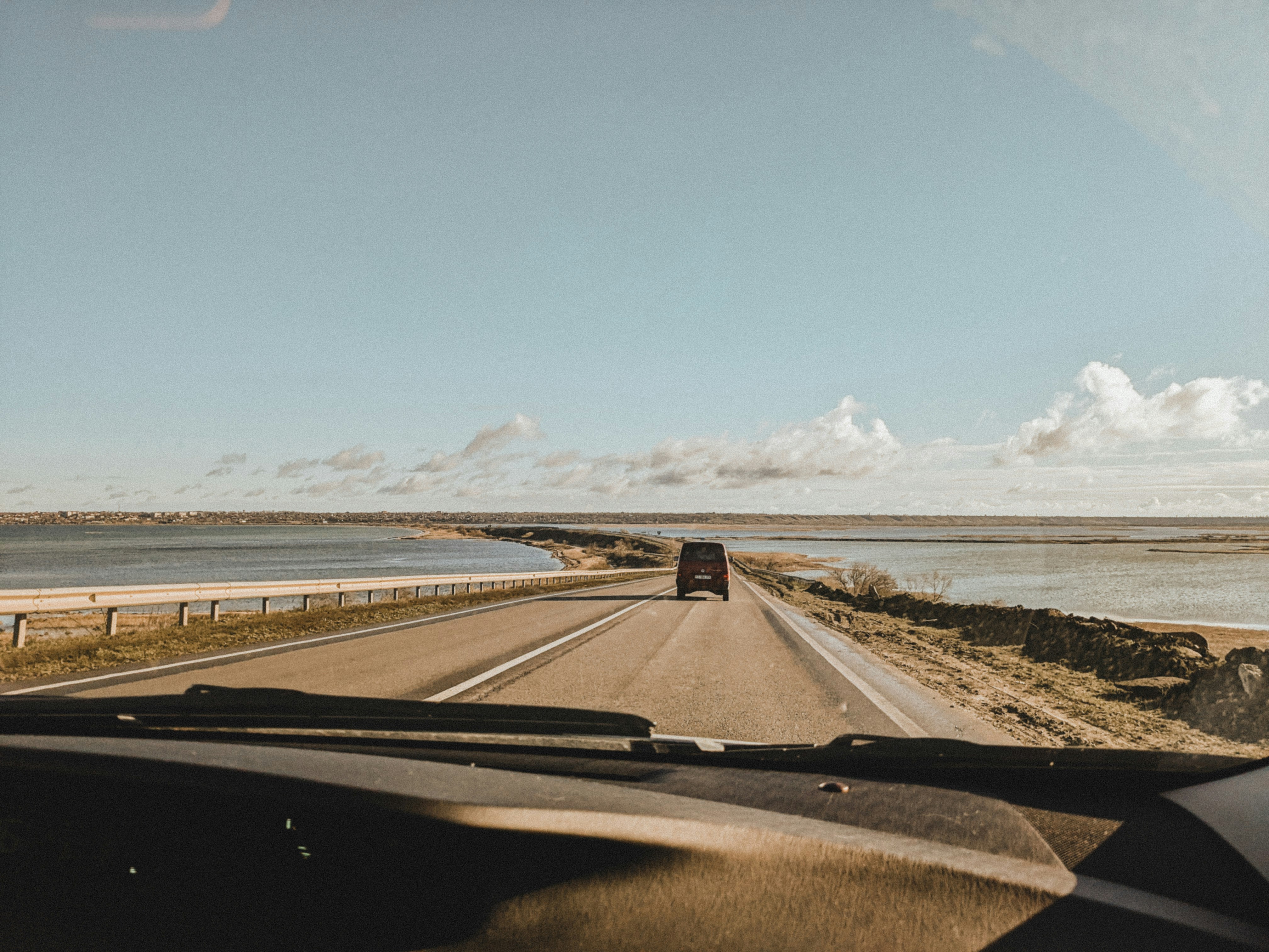 A lone van travels a straight coastal road flanked by salt flats and shallow water beneath a bright sky.