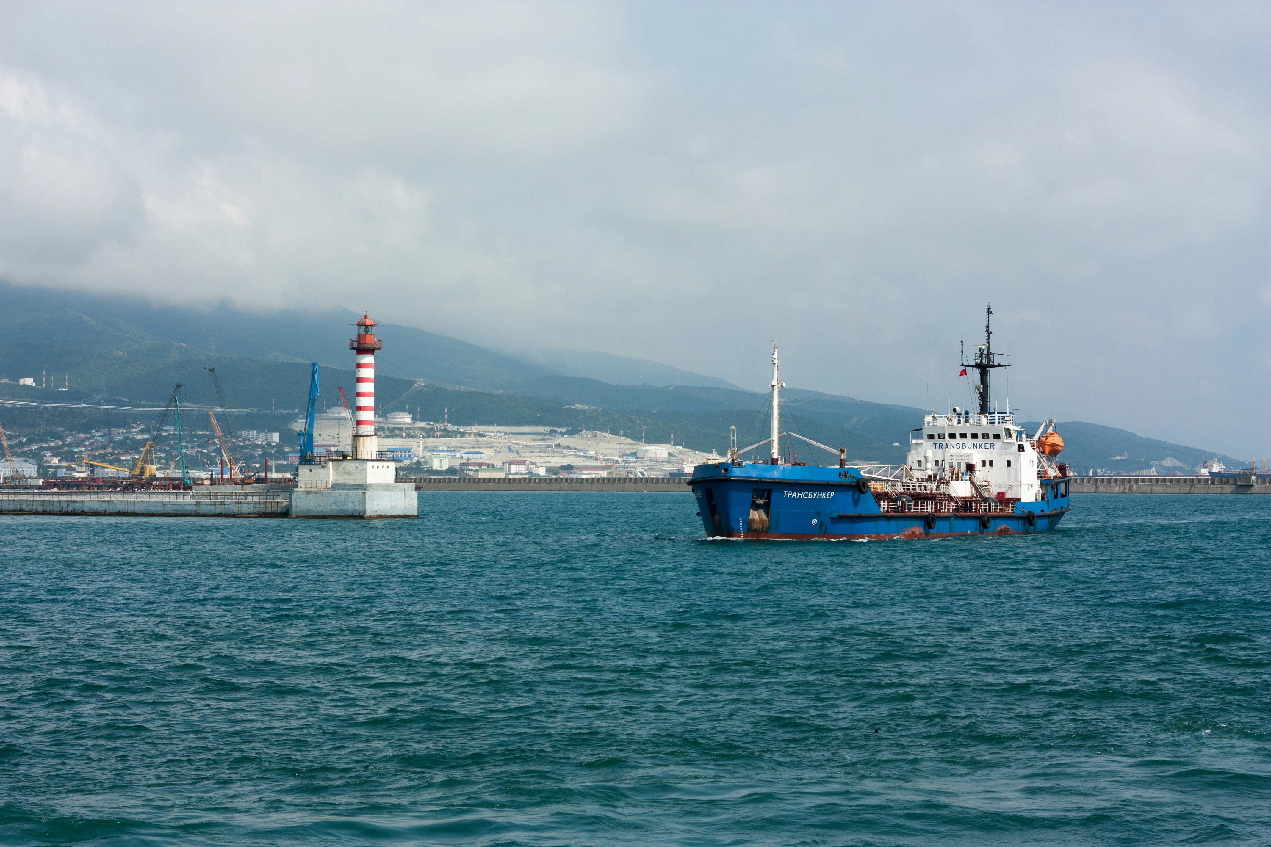 Blue cargo ship sails past a lighthouse under a cloudy sky near a coastal town.