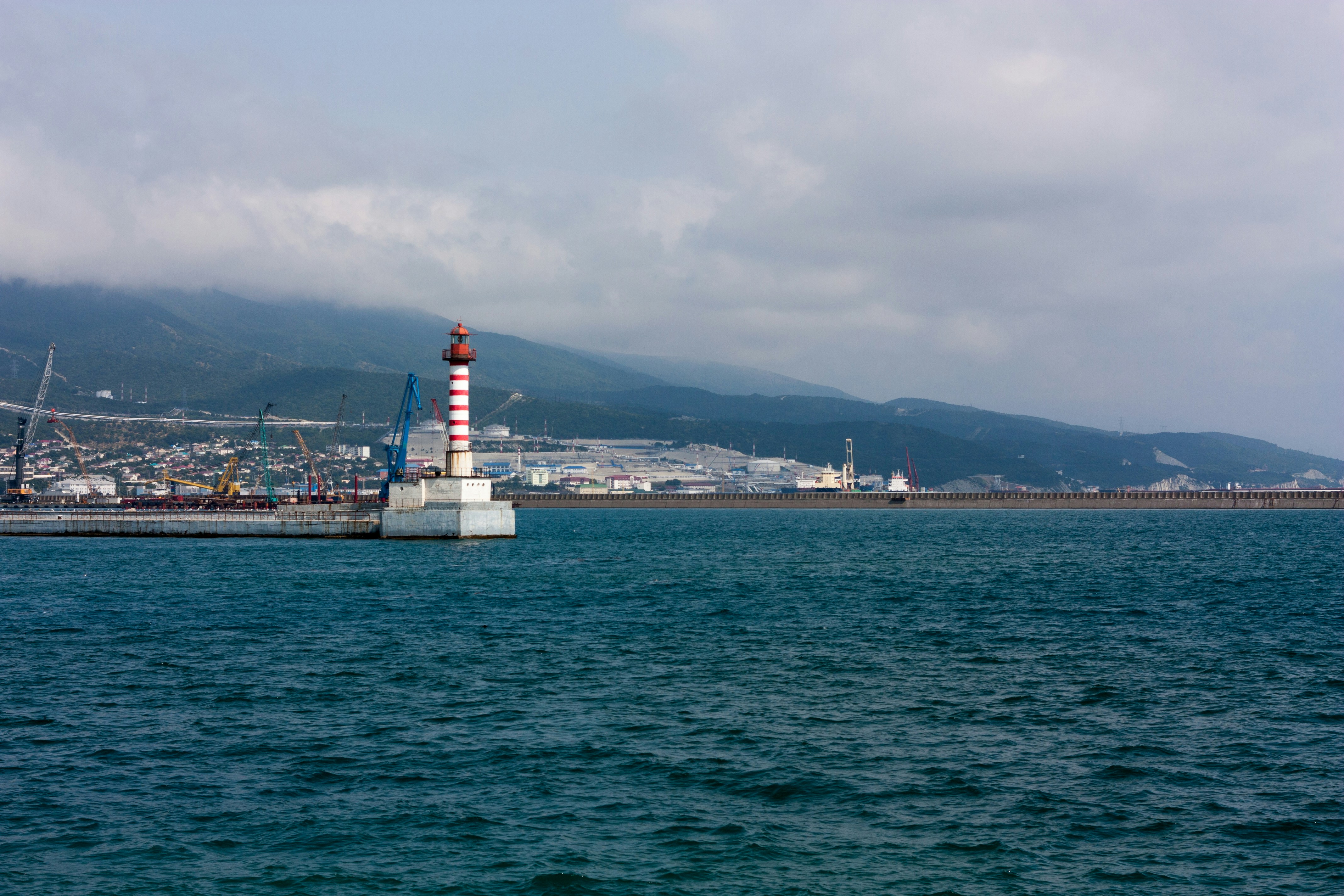 Lighthouse on a pier extends into a calm sea under a cloudy sky, with a mountainous coastline in the background.