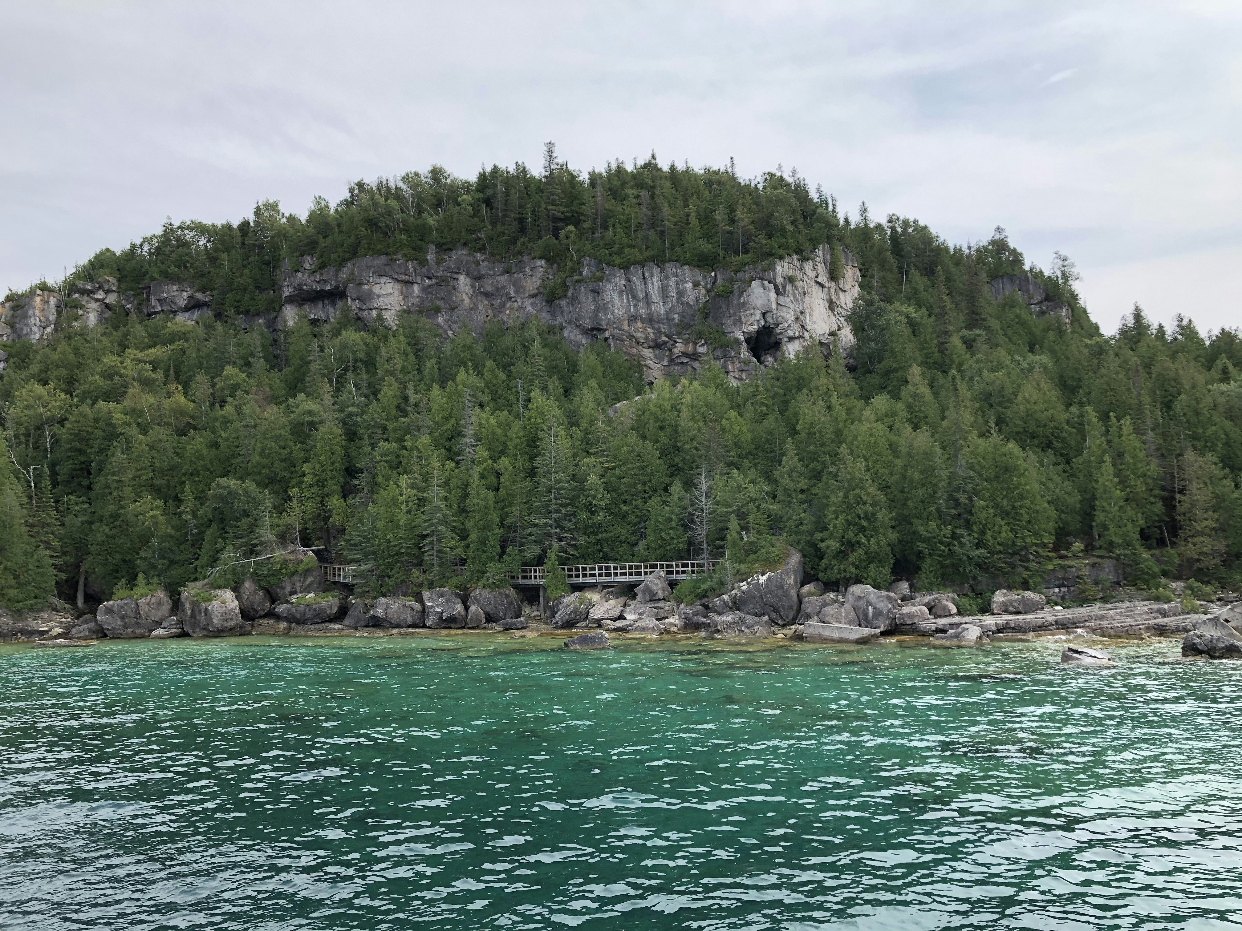 Lush green forest meets rocky shoreline under a cloudy sky, with clear turquoise waters in the foreground. A wooden path leads into the trees.