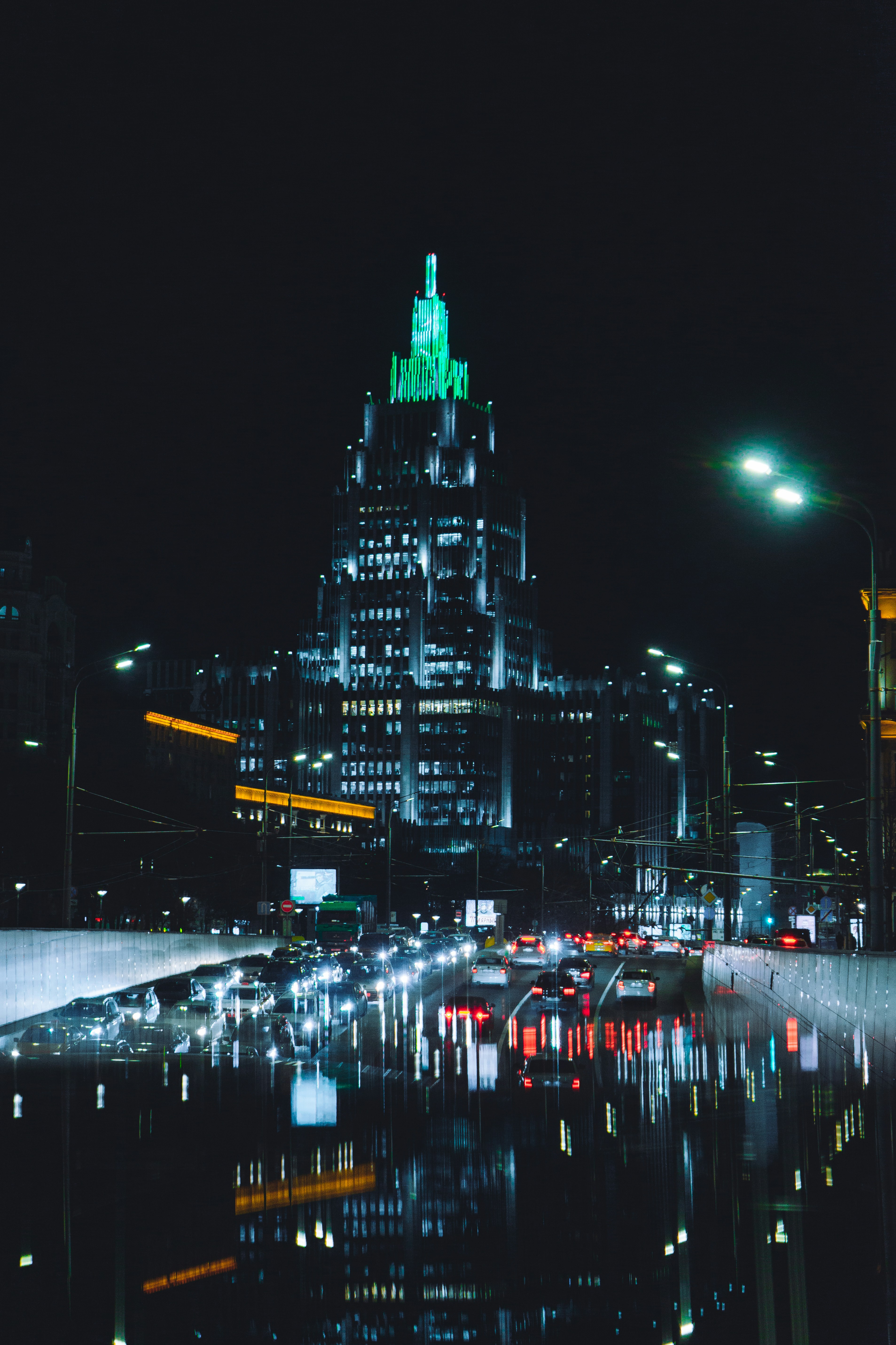 Illuminated skyscraper reflecting in water at night, surrounded by moving traffic. The green beacon atop the building adds a vibrant touch.