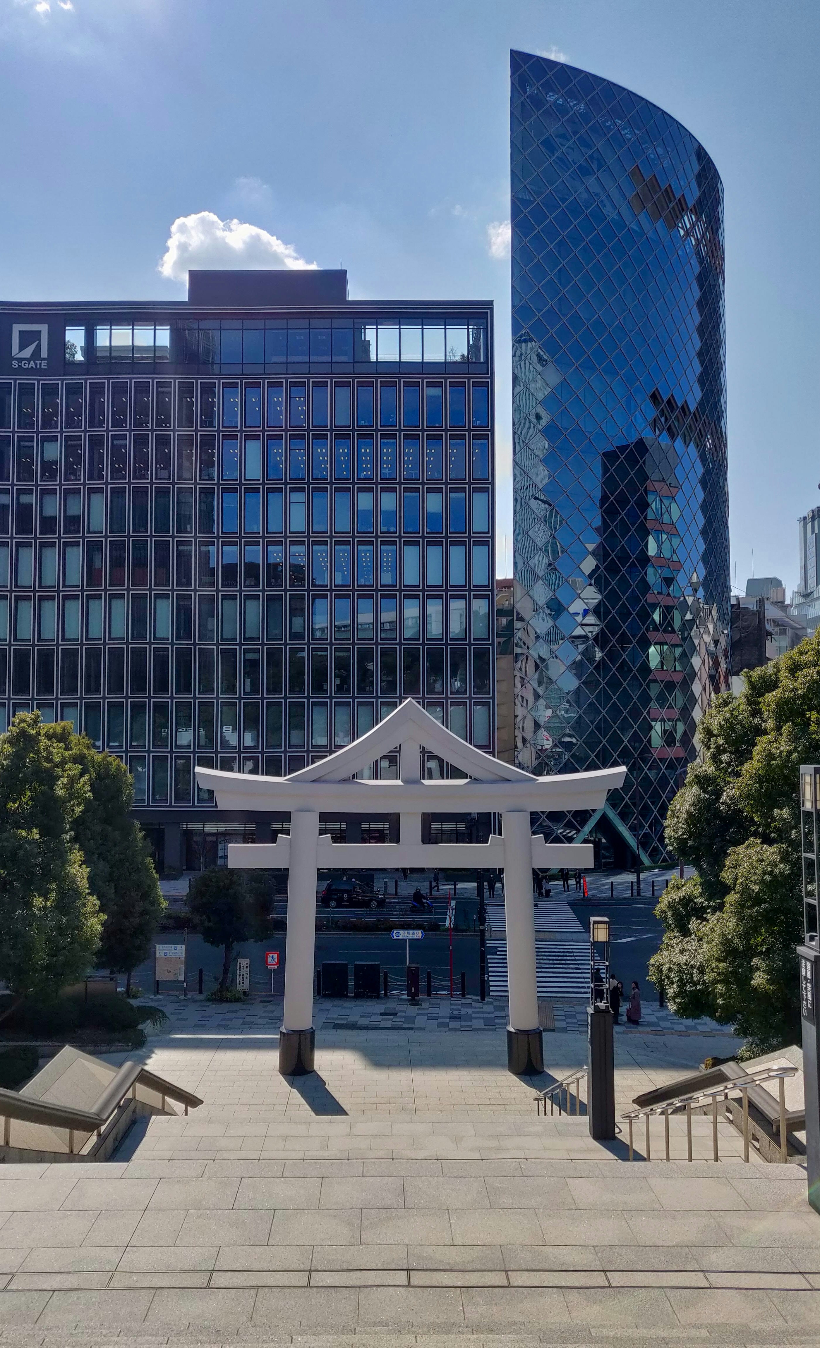Torii gate foreground with modern glass buildings and a curved skyscraper in the background; a civic plaza leads to a corporate complex, capturing a blend of traditional and contemporary architecture.
