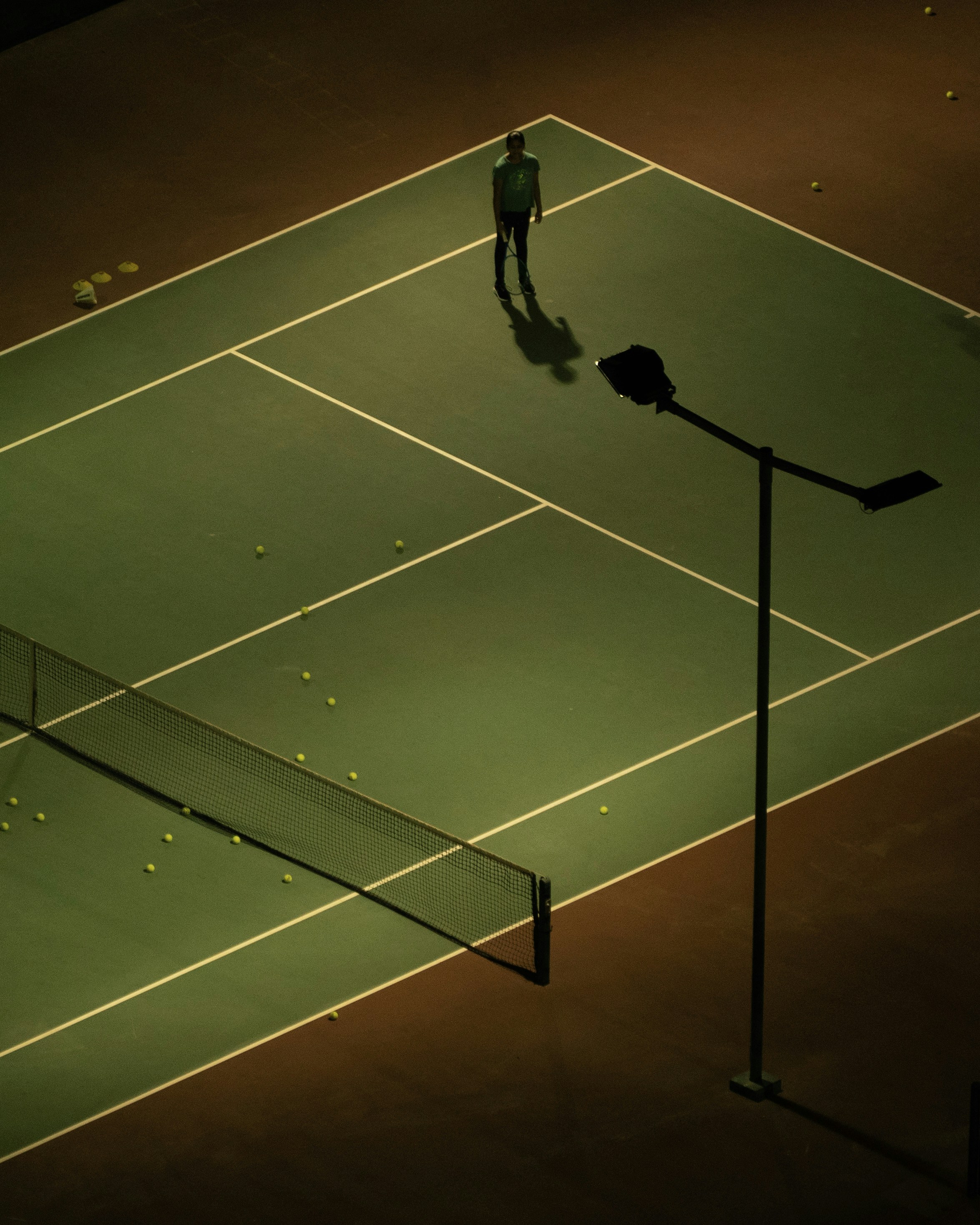 A lone figure stands on a tennis court illuminated by a single light, surrounded by scattered tennis balls. The scene captures the essence of solitude in sports.