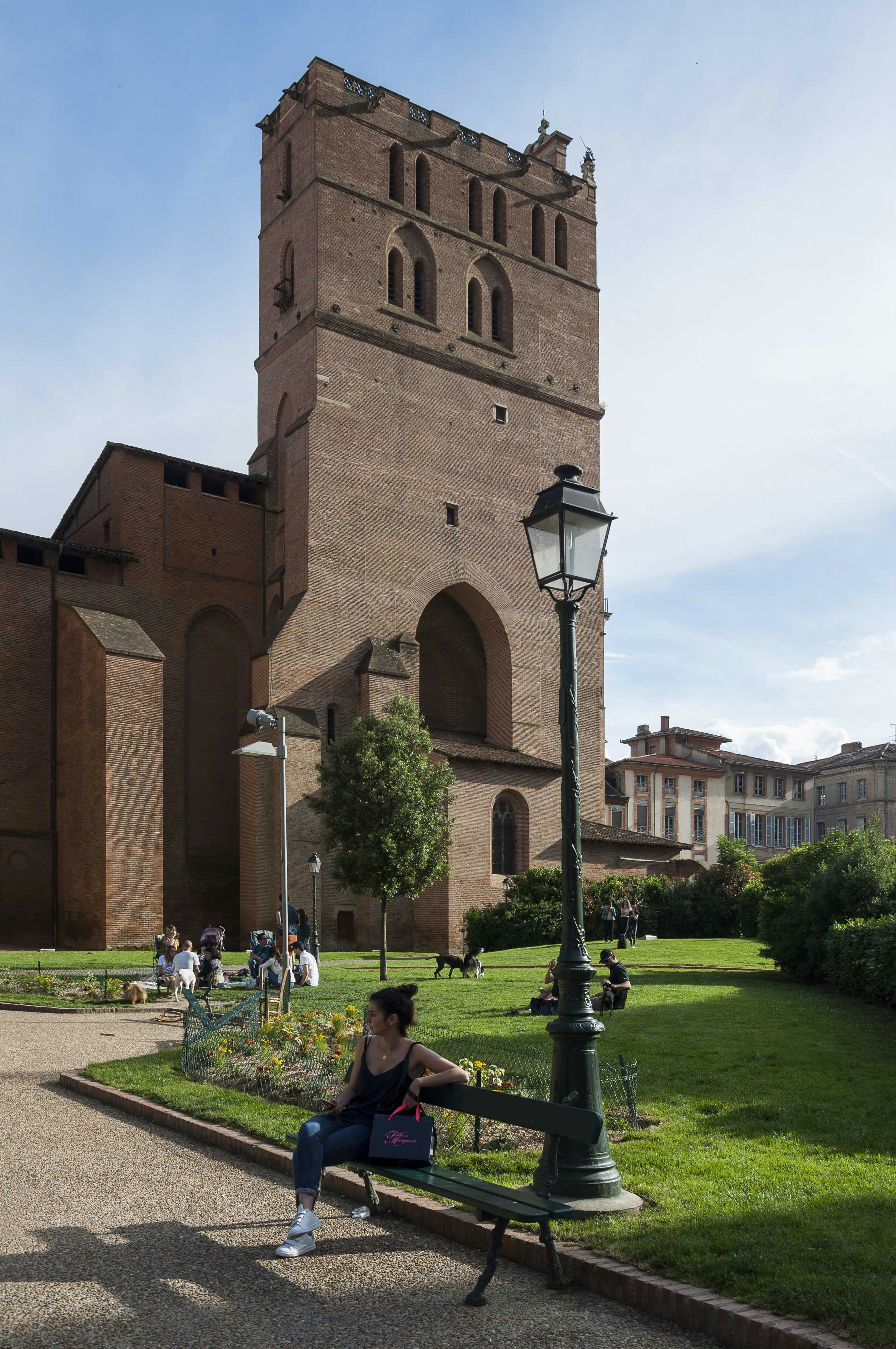 A woman sitting on a bench in a park, with a historic brick tower and lush greenery in the background. The scene captures a blend of architecture and leisure.