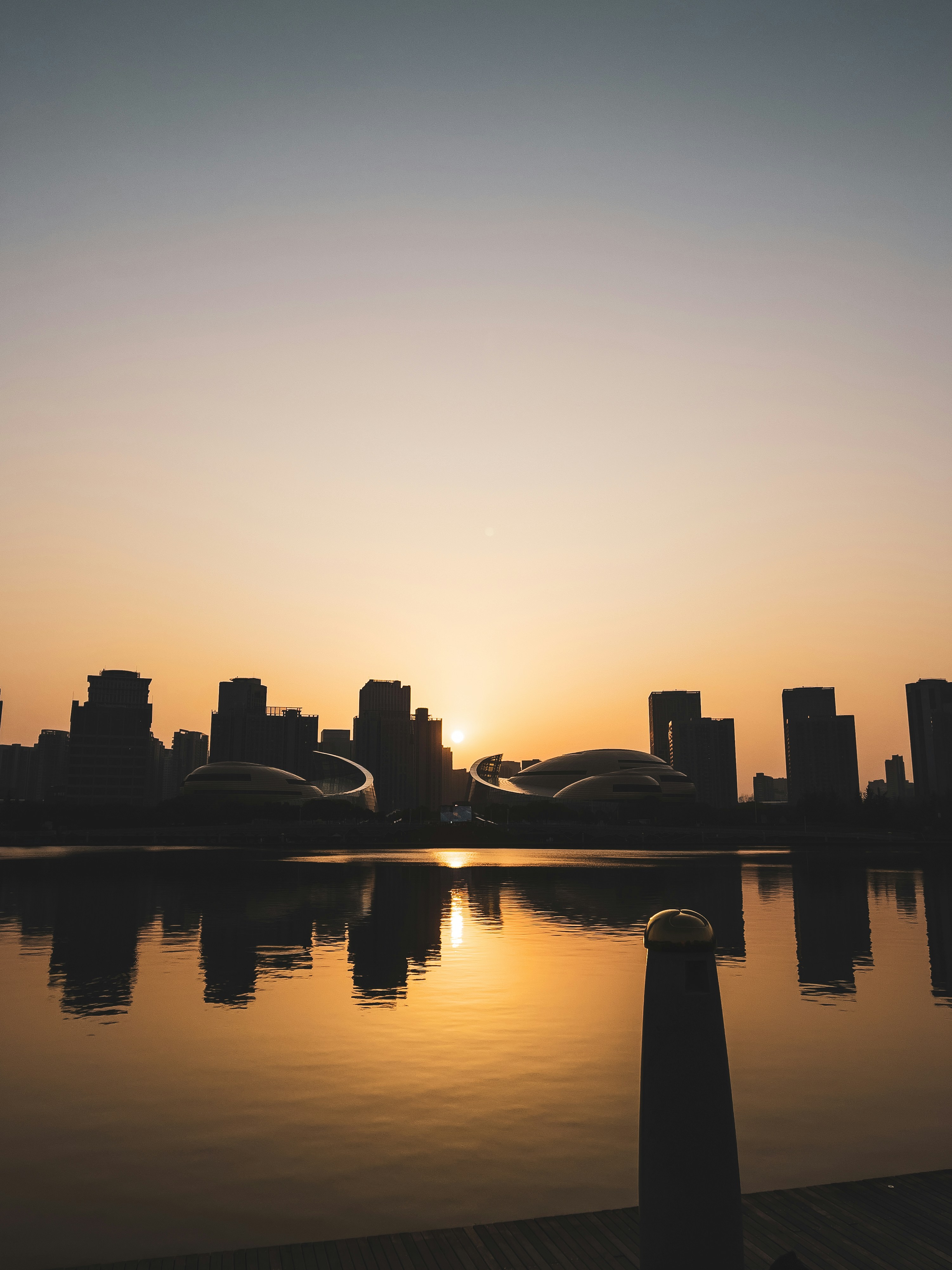 Silhouetted skyline reflecting on water during sunset, with modern architecture and a serene atmosphere.