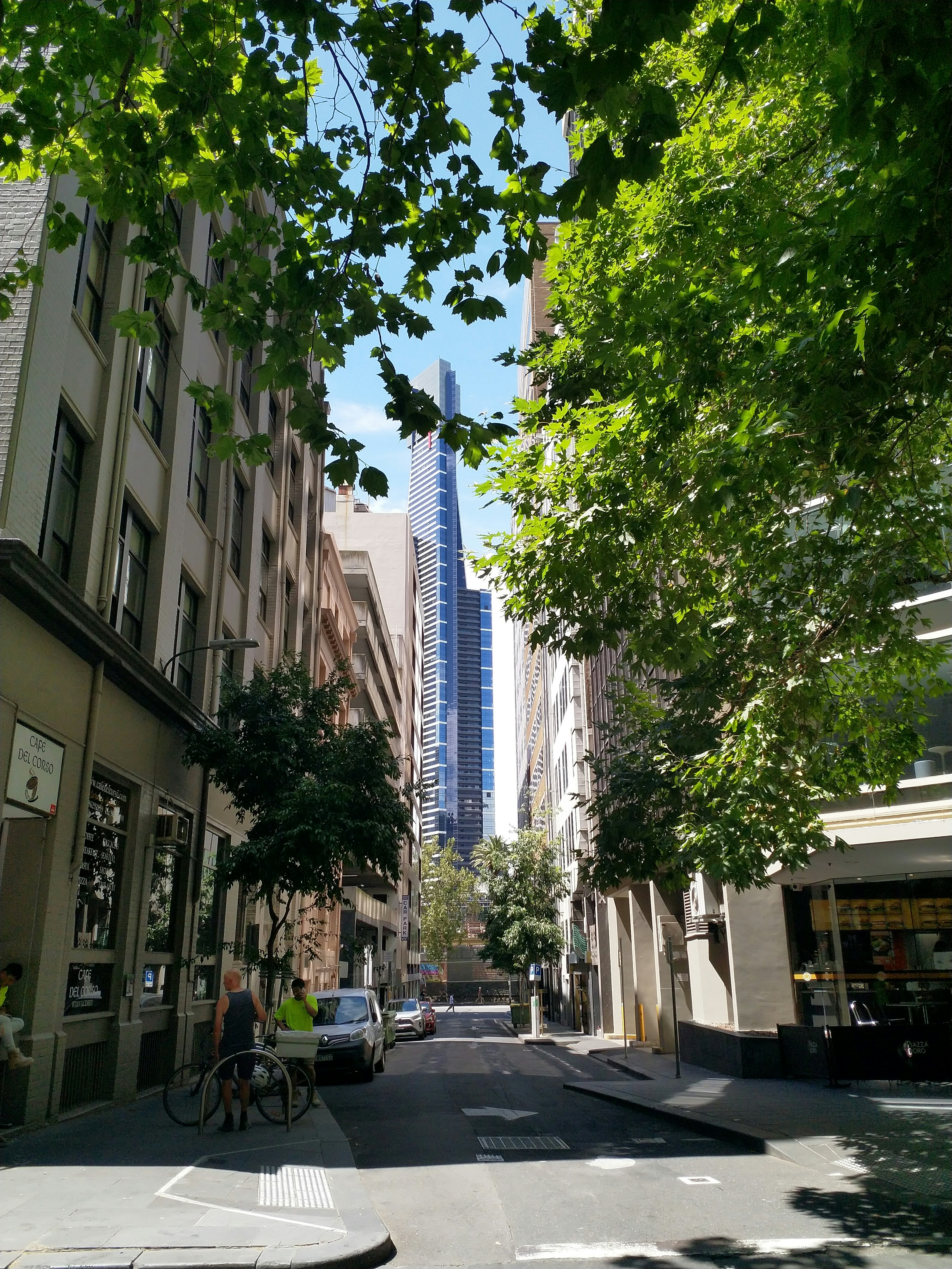 Urban street scene with leafy canopies framing a slender blue glass skyscraper at the center, flanked by historic and modern buildings. A few pedestrians and a cyclist inhabit the sunlit boulevard.