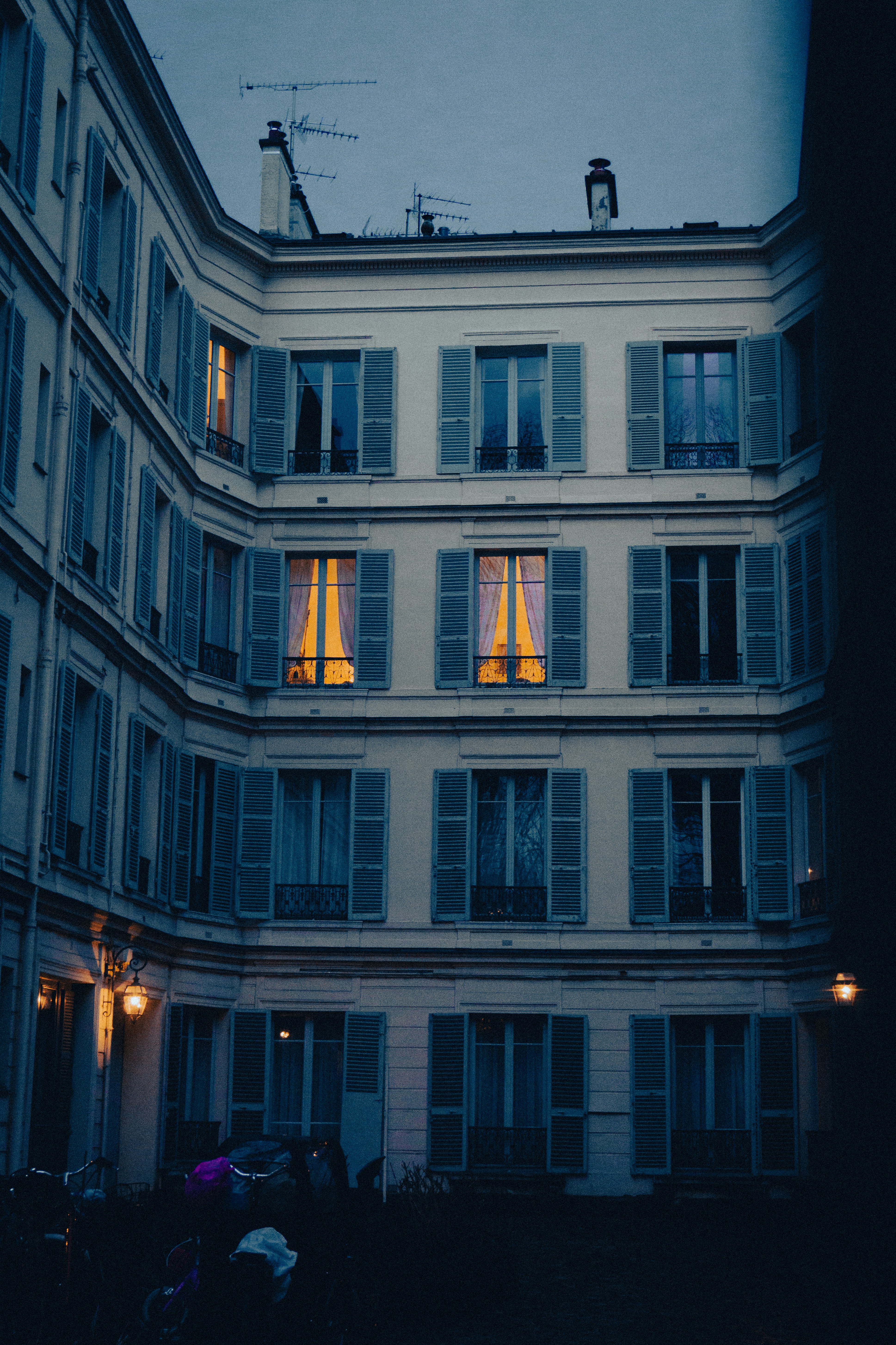 Illuminated windows of a Parisian apartment building reveal glimpses of life inside, framed by classic shutters. The scene evokes a sense of intimacy and warmth amidst the evening chill.