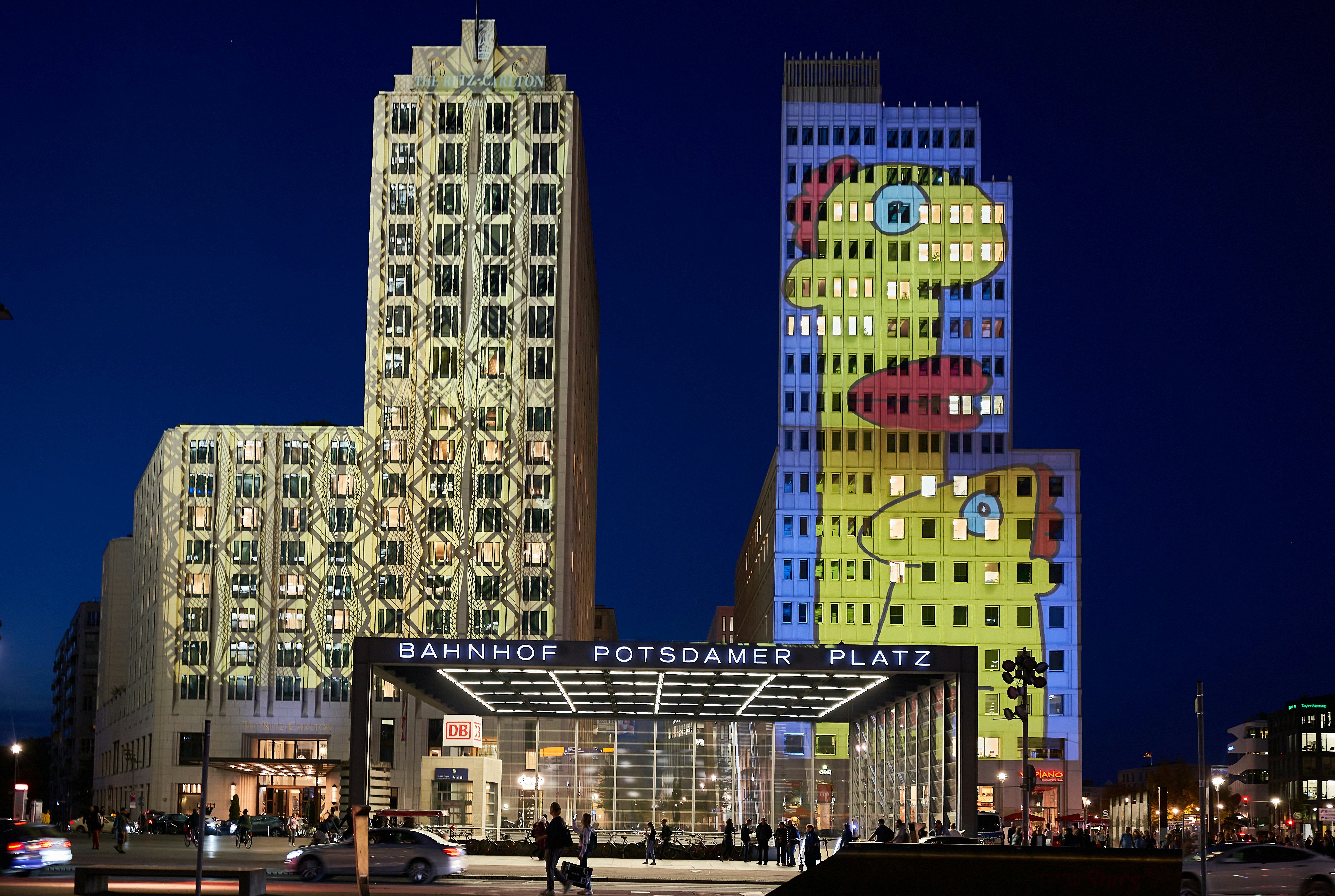 Vibrant projections light up skyscrapers at Potsdamer Platz during nighttime, with a bustling urban scene below.