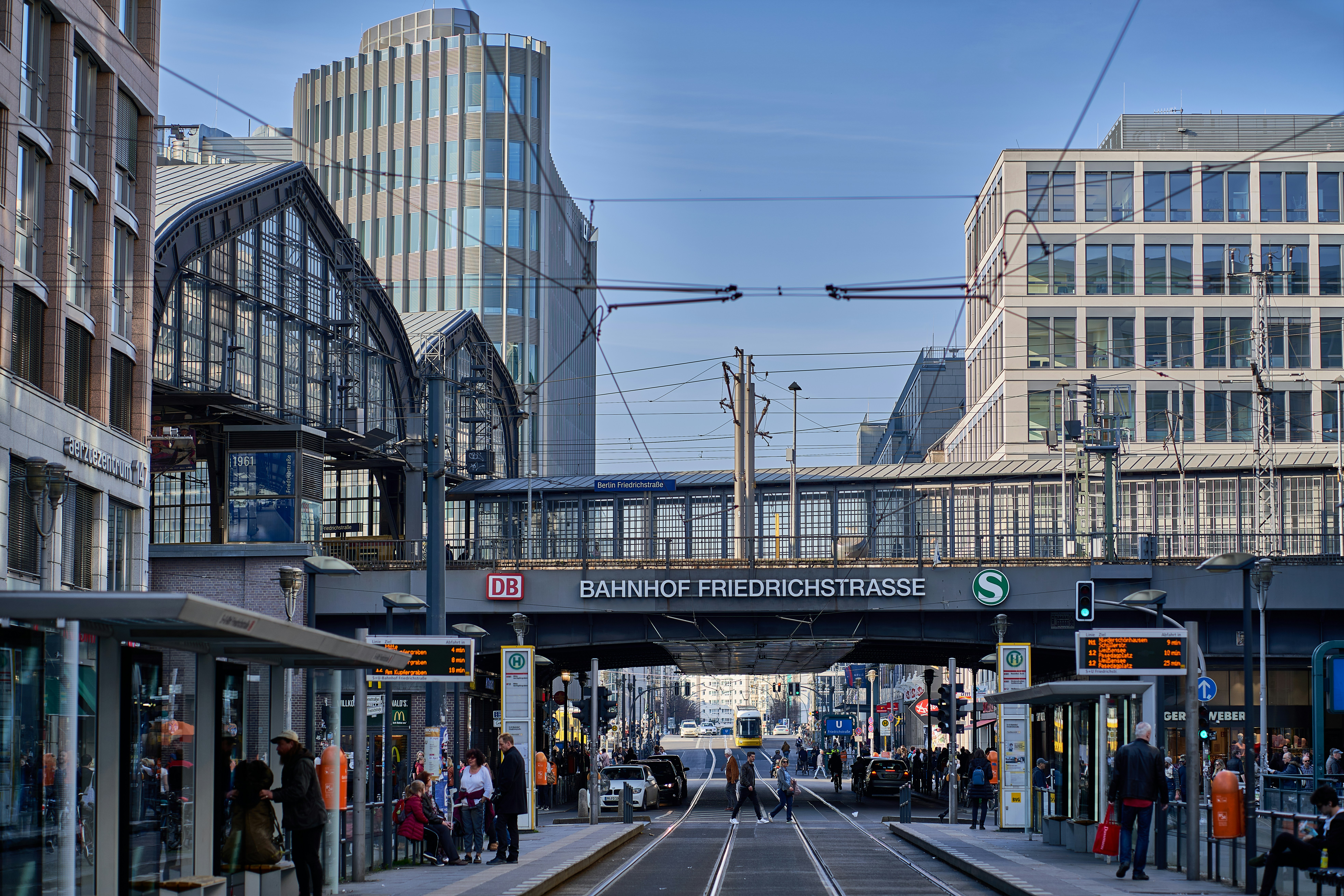 Bustling street scene at Bahnhof Friedrichstrasse, showcasing modern architecture and urban life. The tramway runs through the center, flanked by commuters and shops.