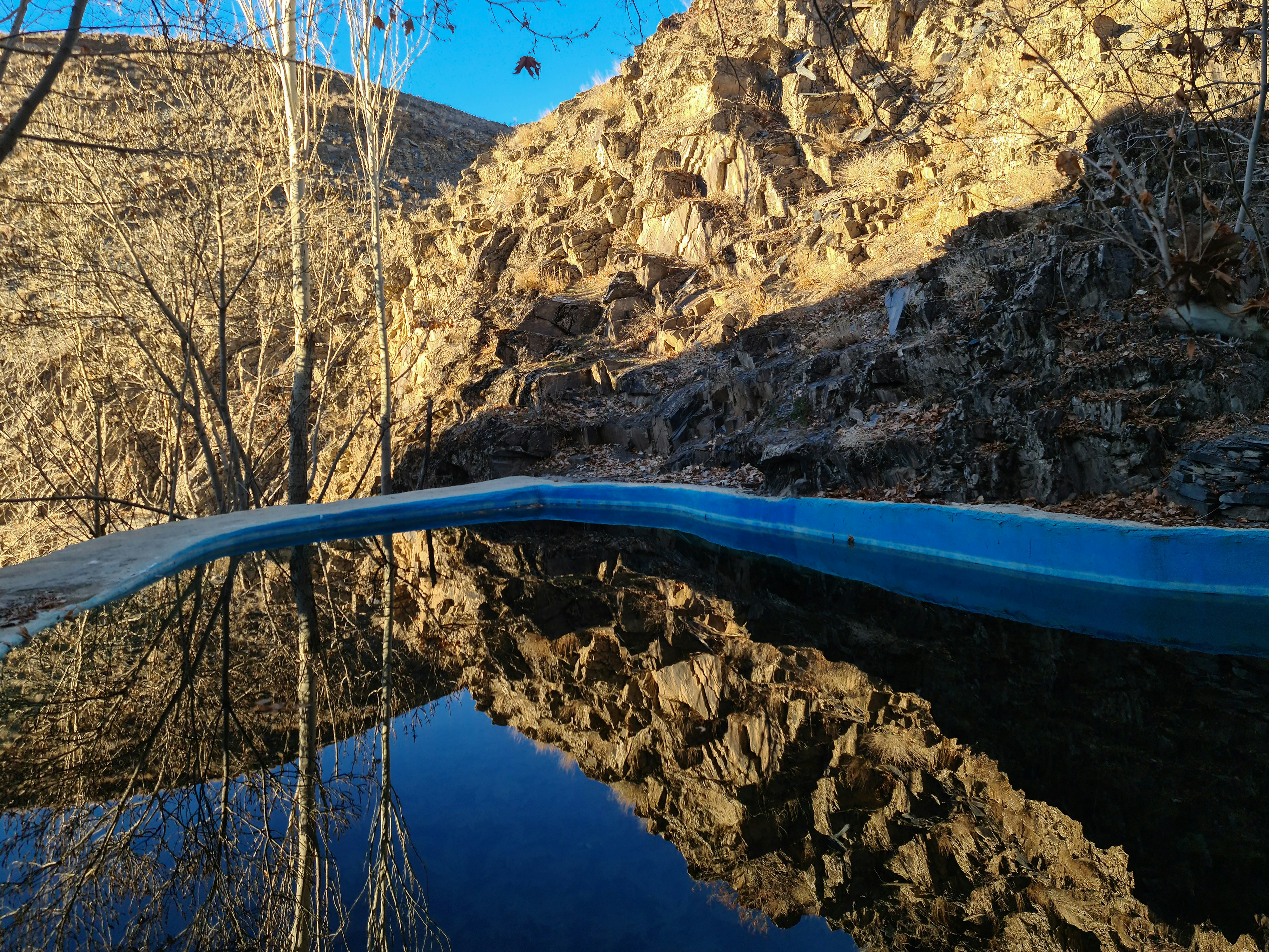 Still water reflecting rocky terrain and bare trees under a clear blue sky.
