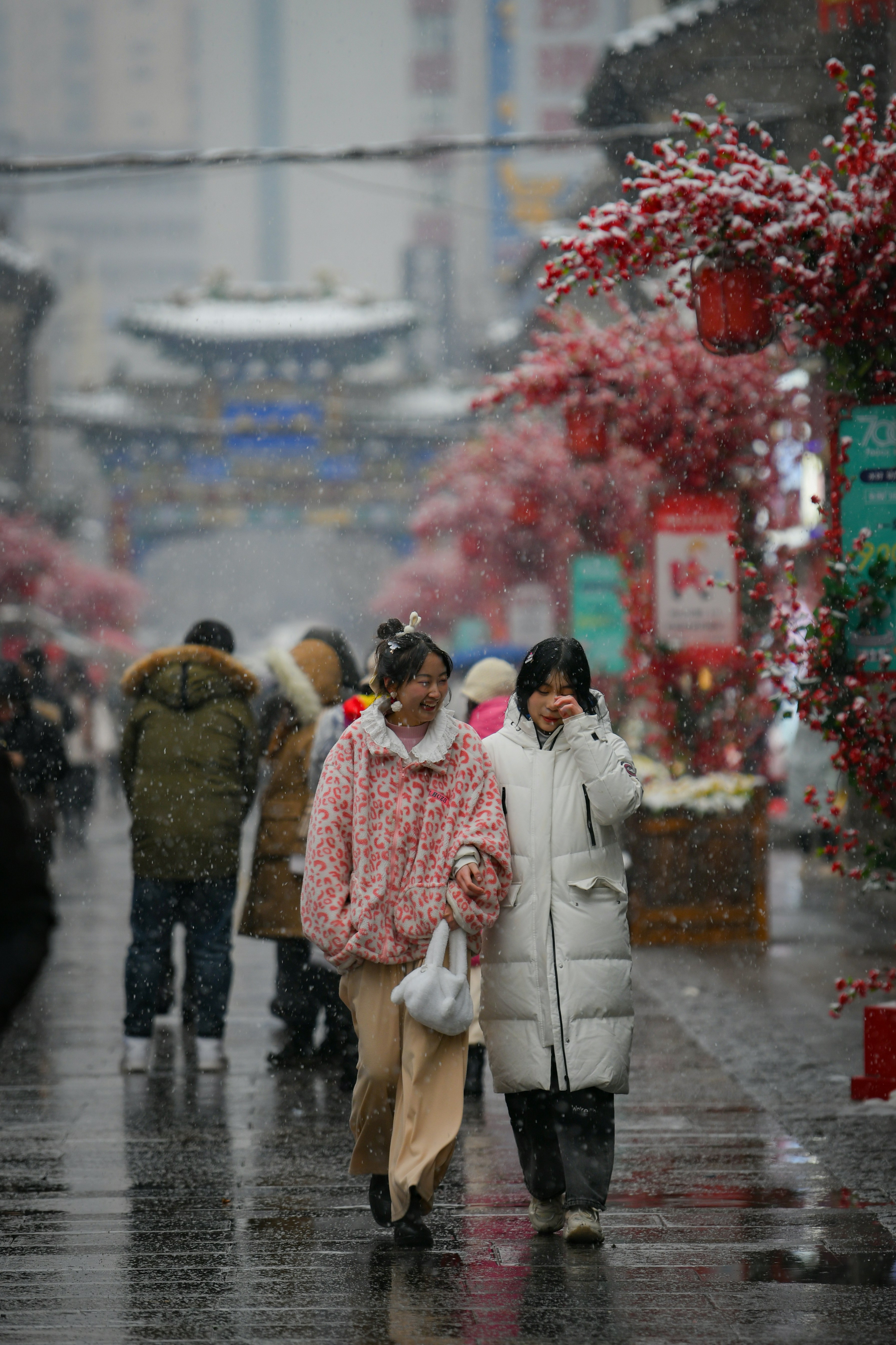 Two women in cozy winter attire walk side by side along a snow-covered street adorned with vibrant decorations. The scene captures a joyful moment amidst a flurry of snowflakes.
