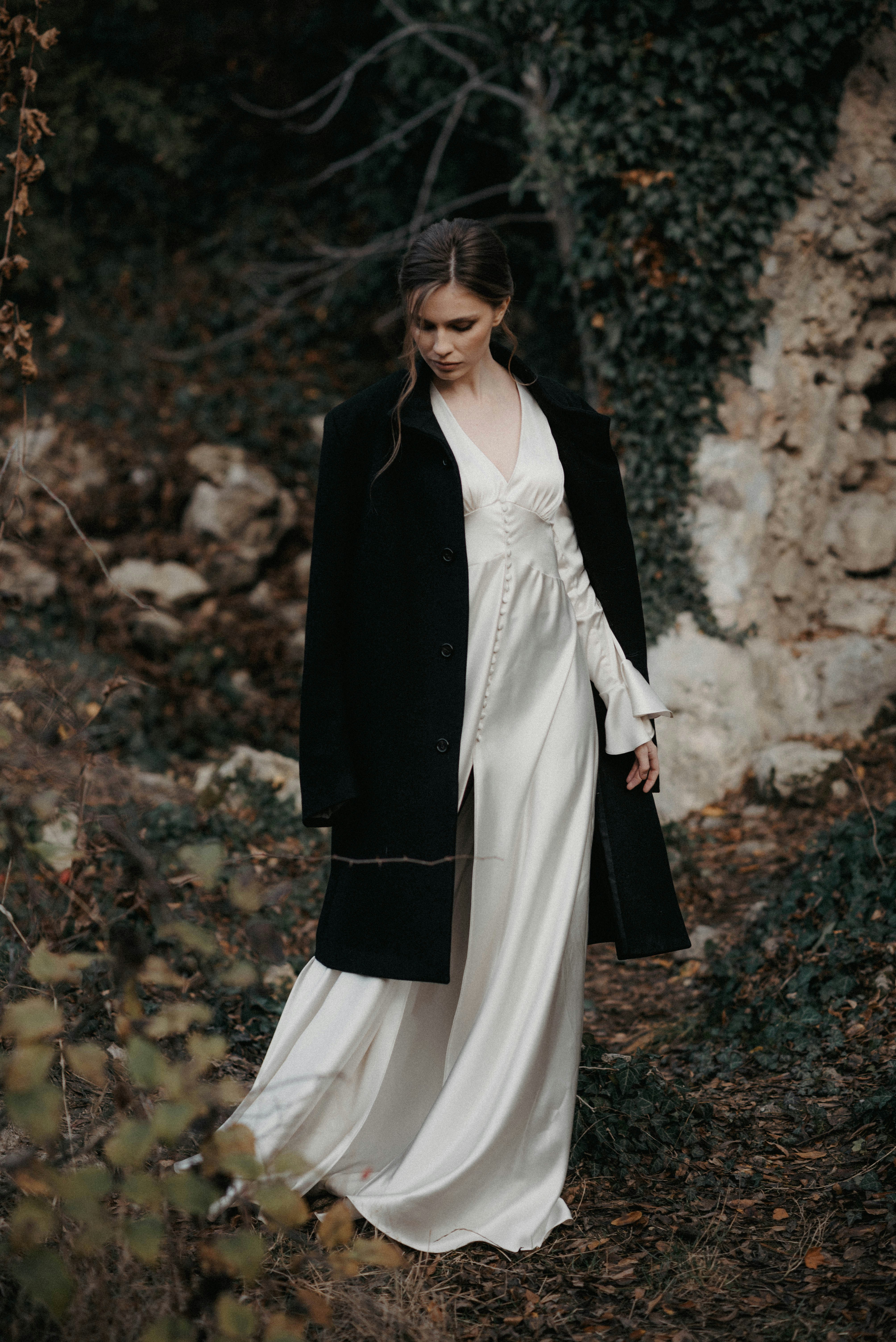 Model in a flowing white gown and black coat stands thoughtfully in a wooded setting, surrounded by earthy tones and textured foliage.