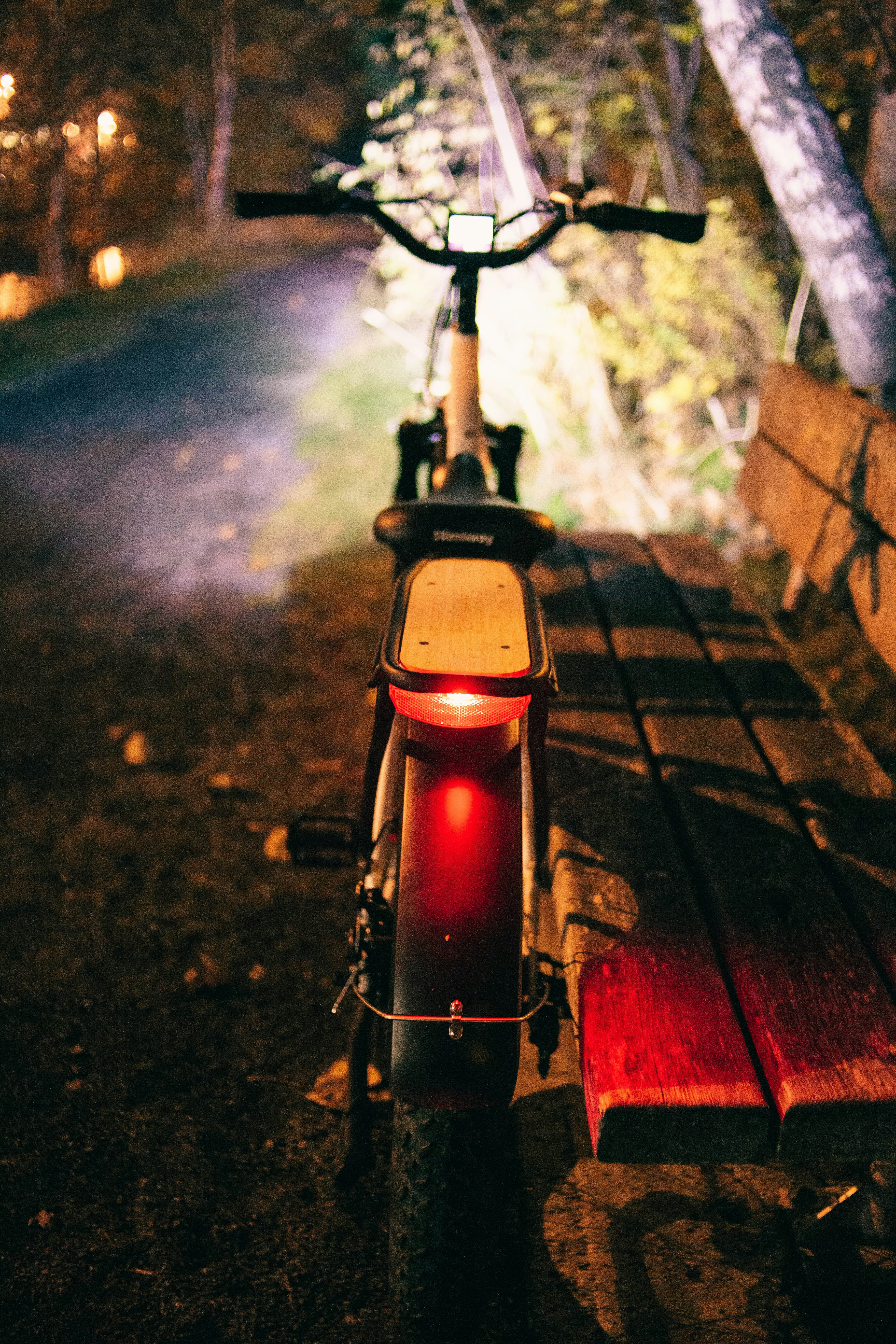 Rear view of a bicycle parked beside a wooden bench on a dimly lit path, showcasing the bike's rear light and the surrounding nature. 