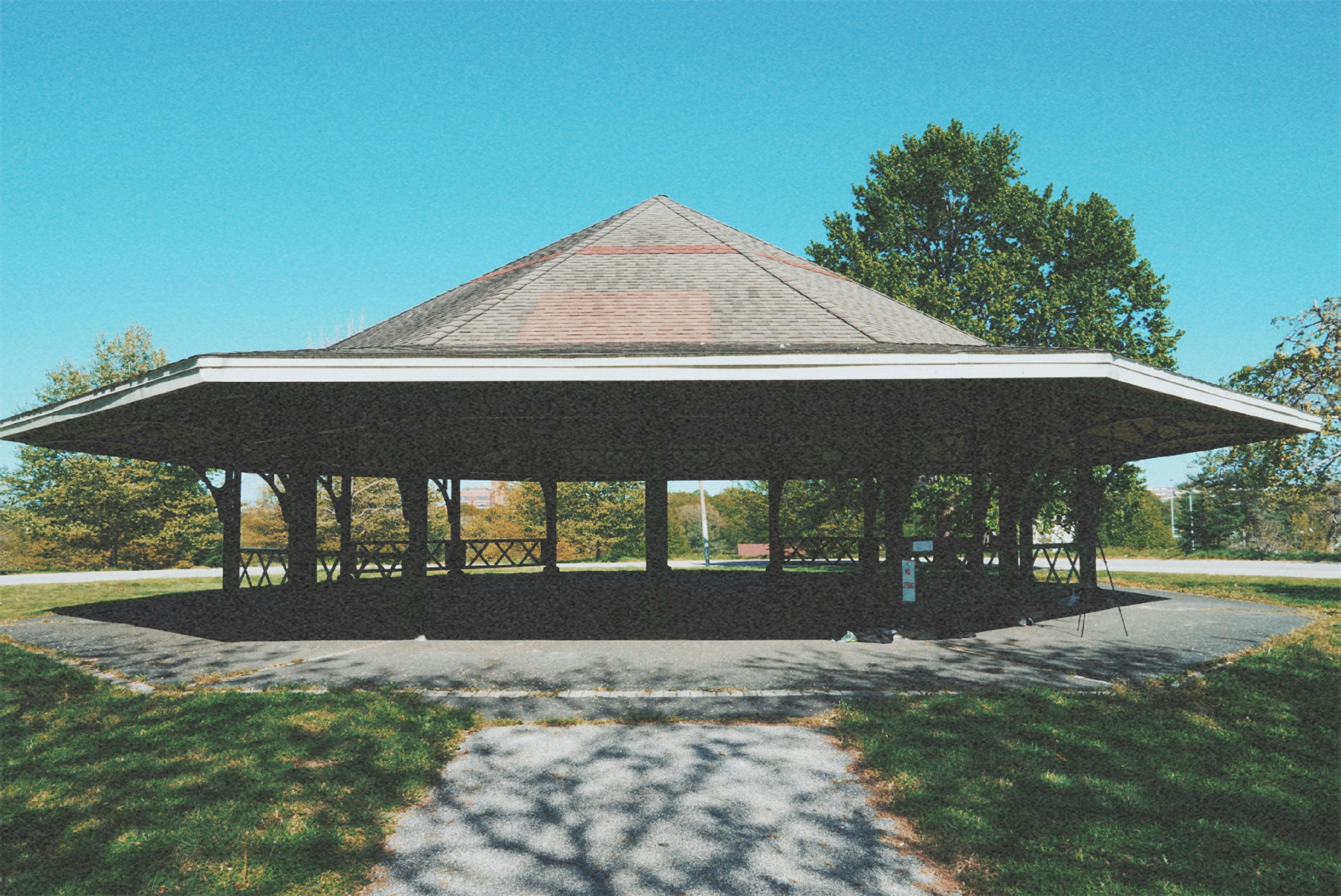 Open-sided gazebo centered in a sunlit park with surrounding trees and a clear sky.