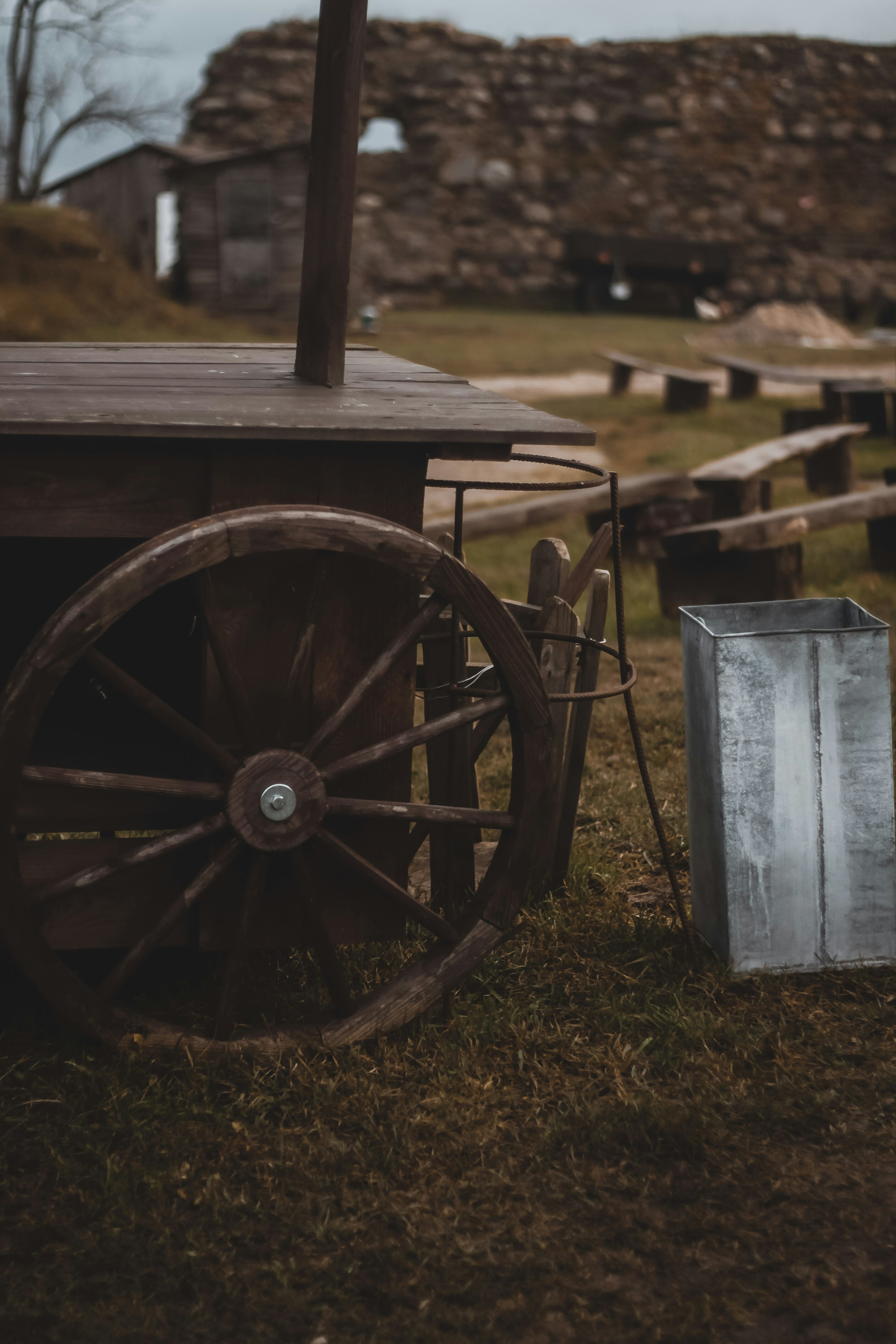 Weathered wooden cart wheel beside a metal bucket in a serene outdoor setting, hinting at a bygone era. Ruins of stone structures loom in the background.