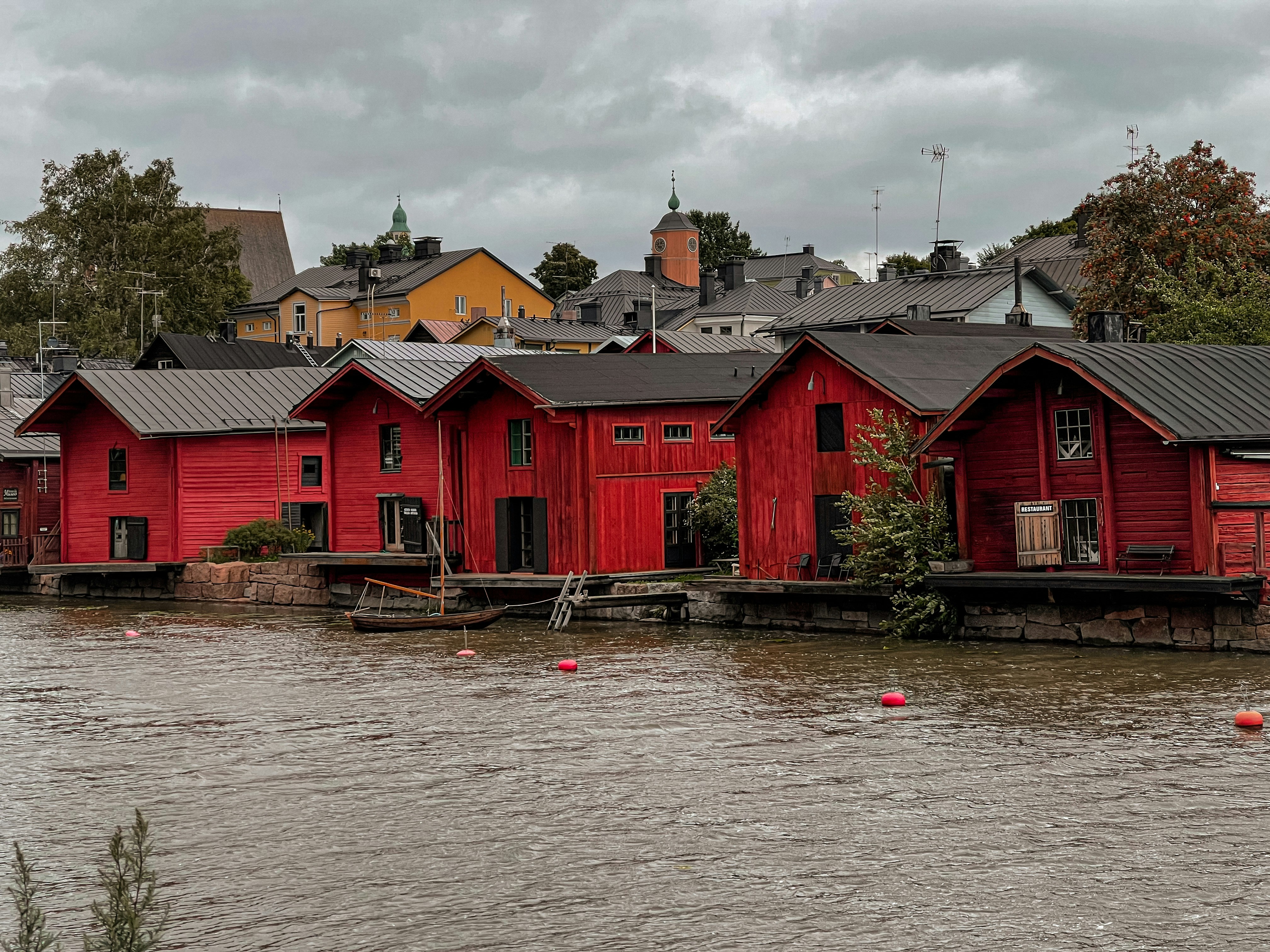 Vibrant red wooden buildings line the edge of a calm river under a cloudy sky.