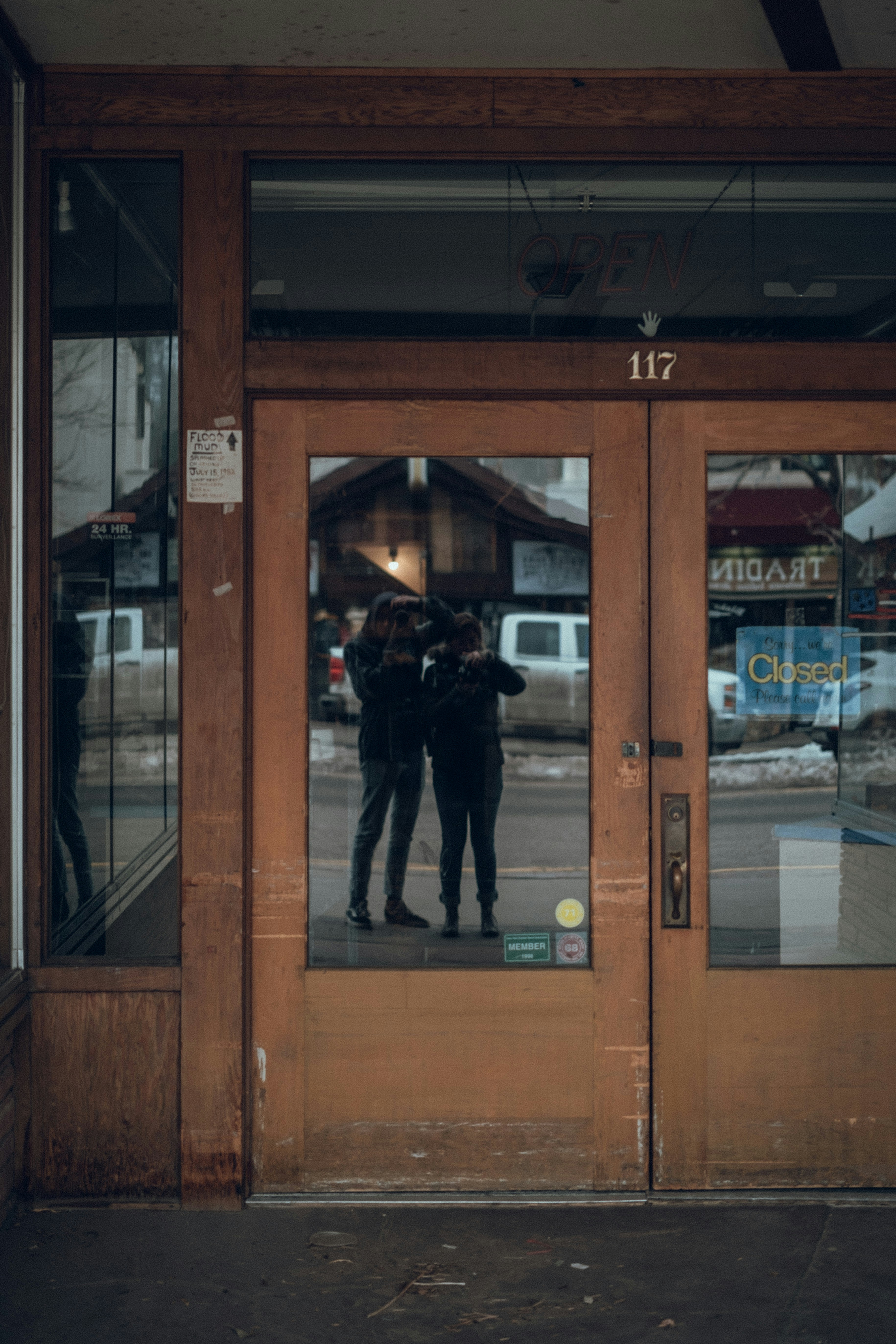 Wooden storefront with reflective glass displaying a 'Closed' sign, capturing two figures in the reflection. The ambiance hints at a calm, overcast day.