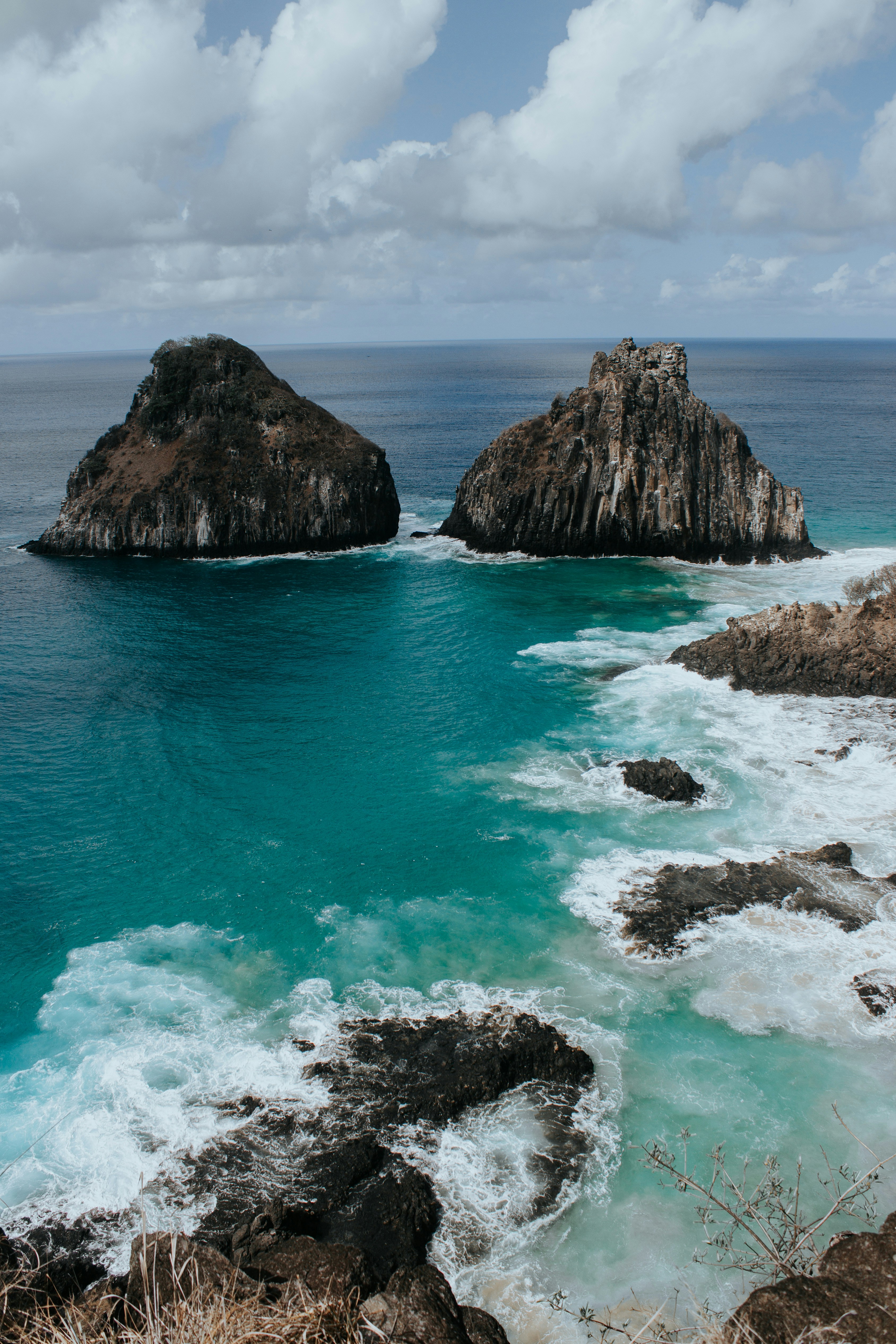 Two rocky islands rise from vibrant turquoise waters, surrounded by frothy waves and a rugged coastline.