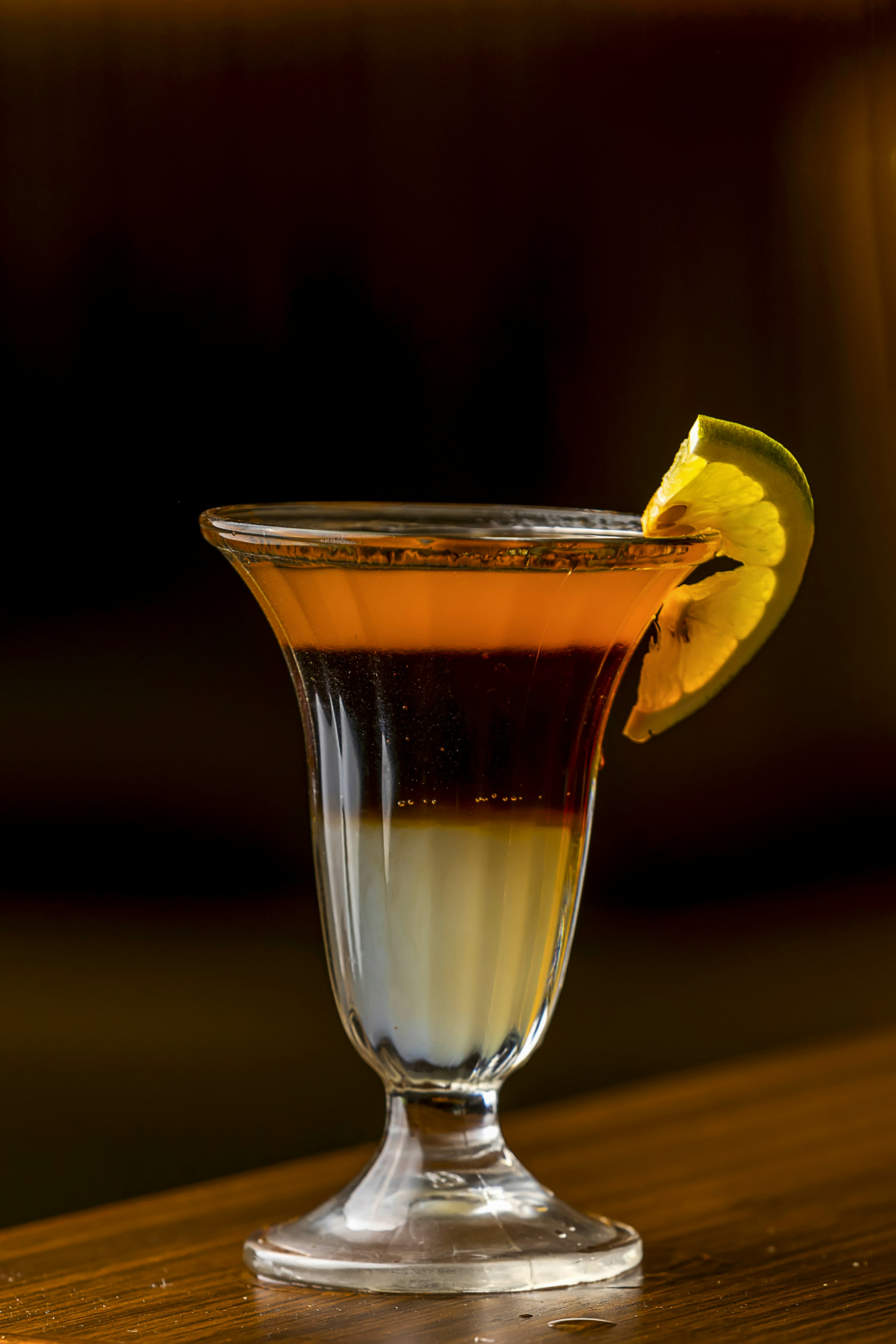 A beautifully layered cocktail served in a decorative glass, garnished with a slice of orange. The contrasting colors create an inviting visual appeal.