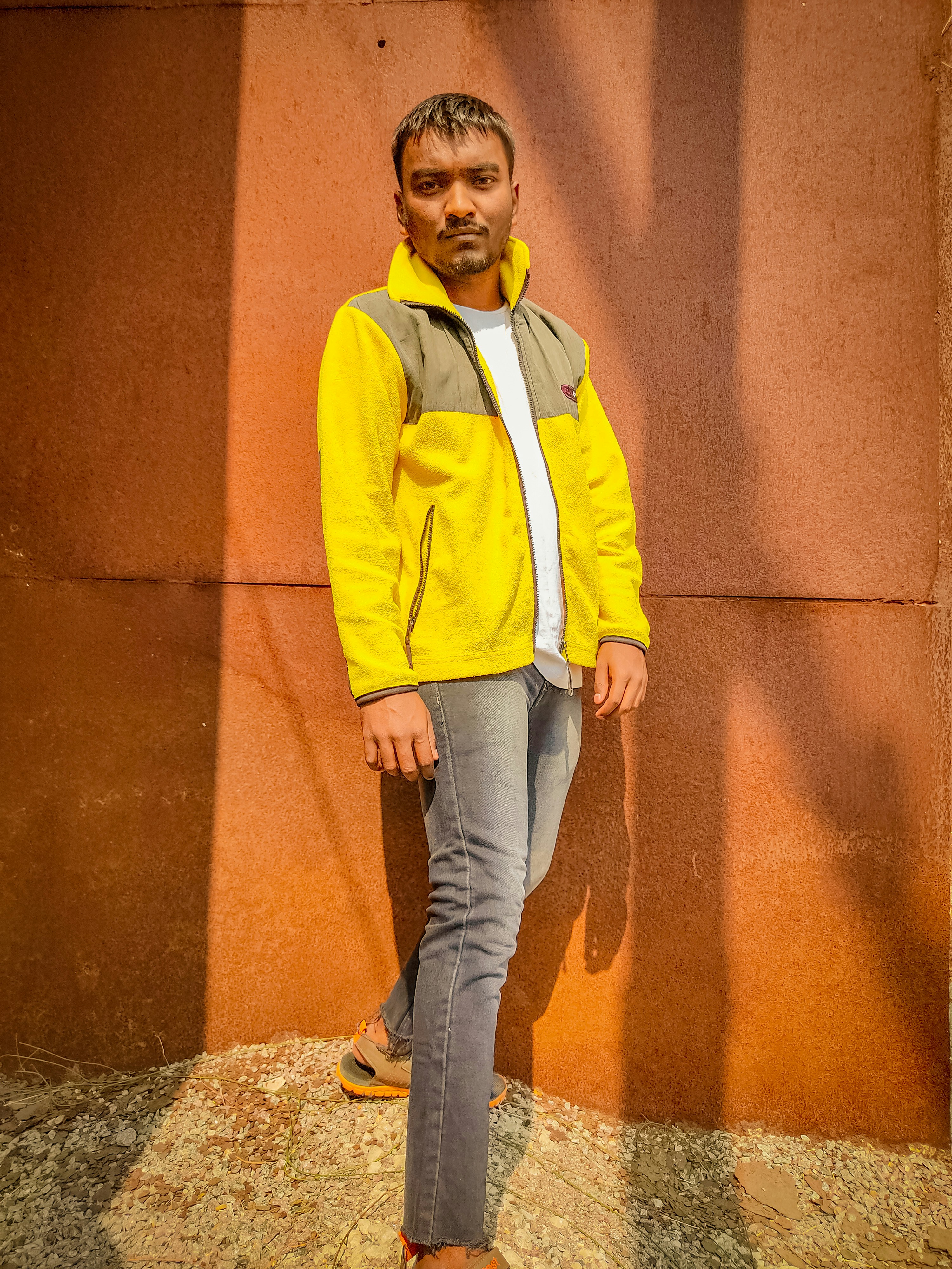 A young man in a vibrant yellow jacket poses confidently against a textured brown backdrop, with shadows creating a dynamic interplay of light and color.