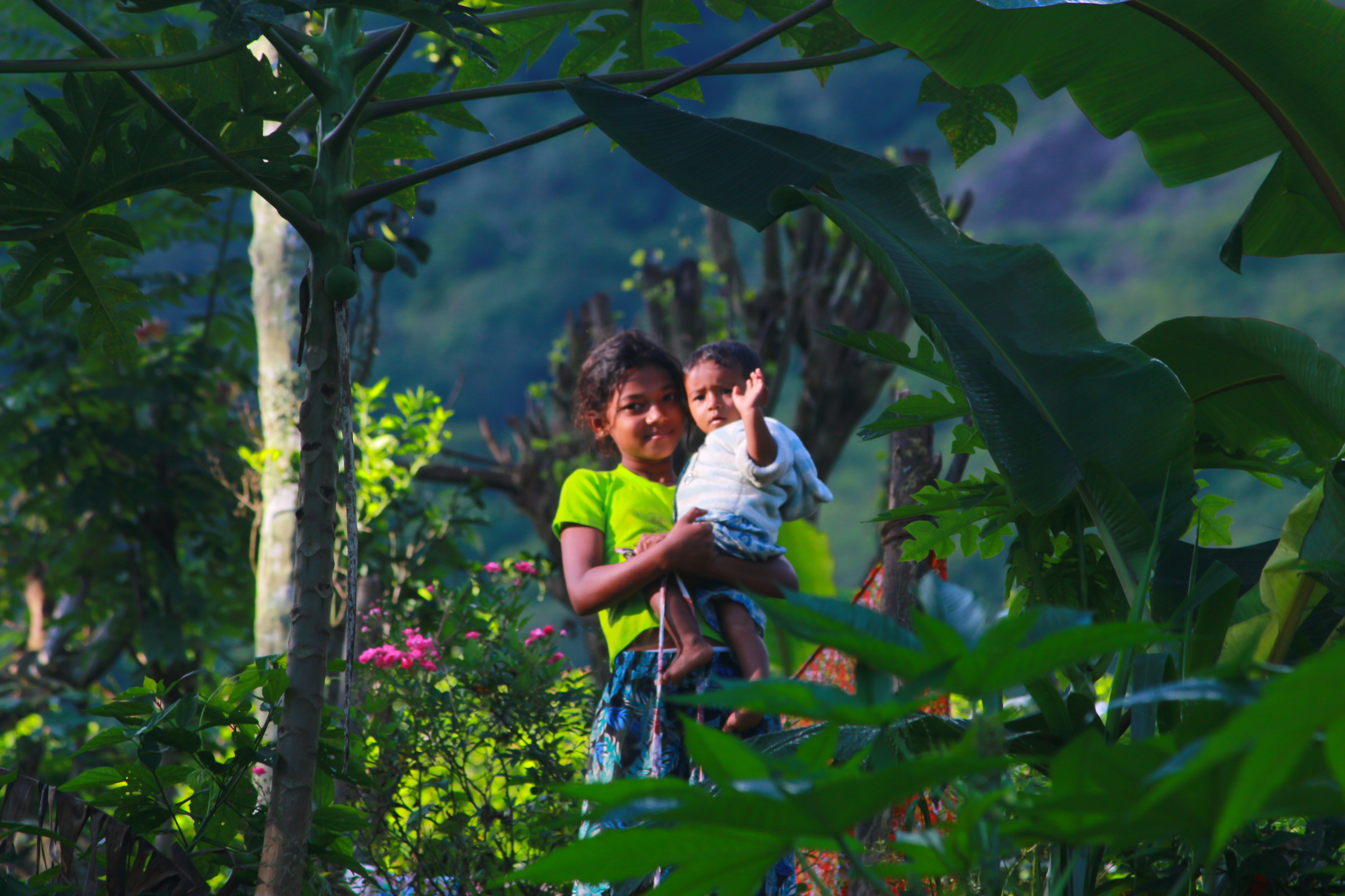 Young girl holding a baby amidst lush greenery and vibrant flowers, capturing a moment of tenderness in a natural setting.