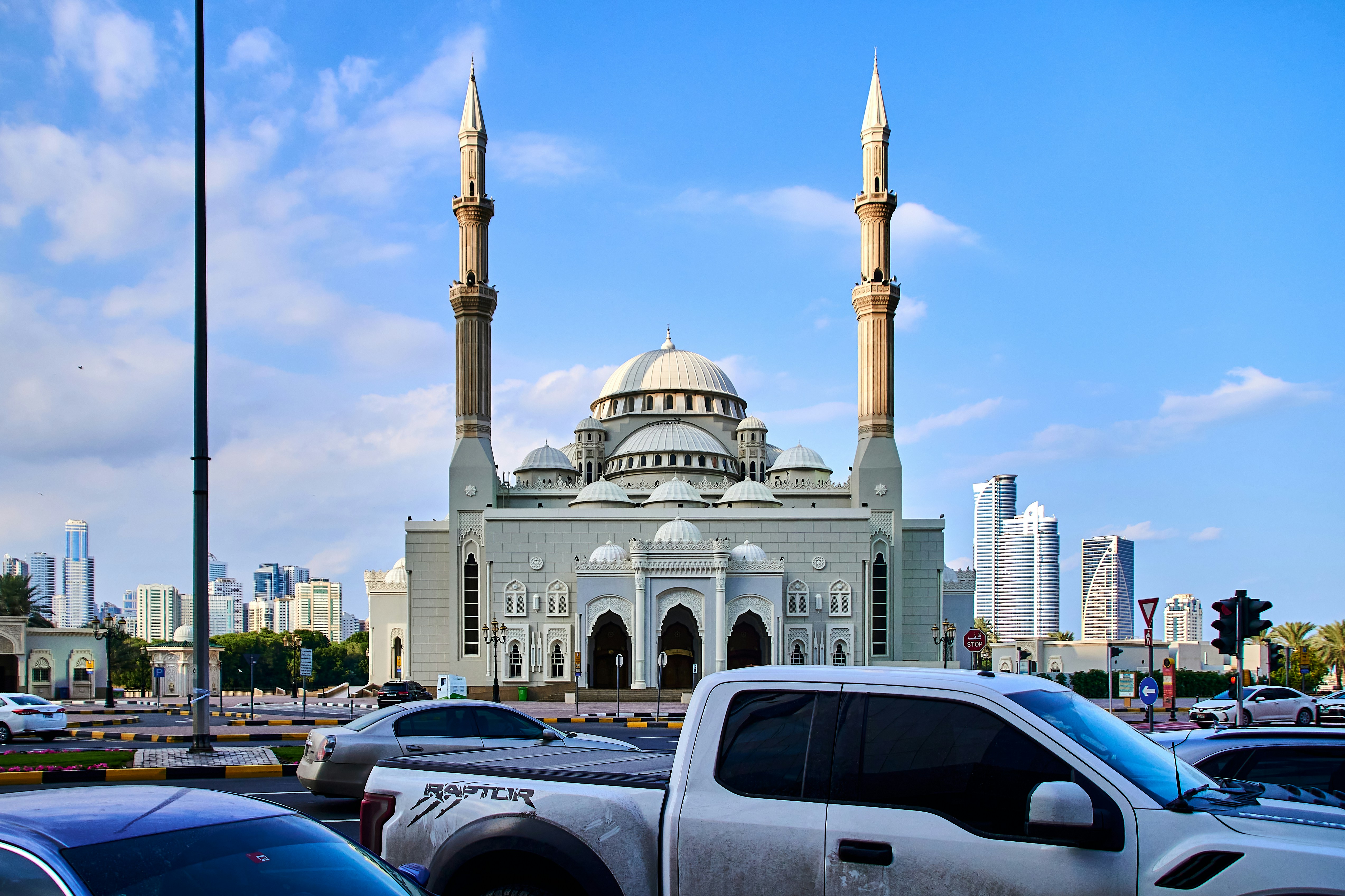 Grand mosque with intricate design and towering minarets set against a city skyline. Traffic in the foreground adds a dynamic element.