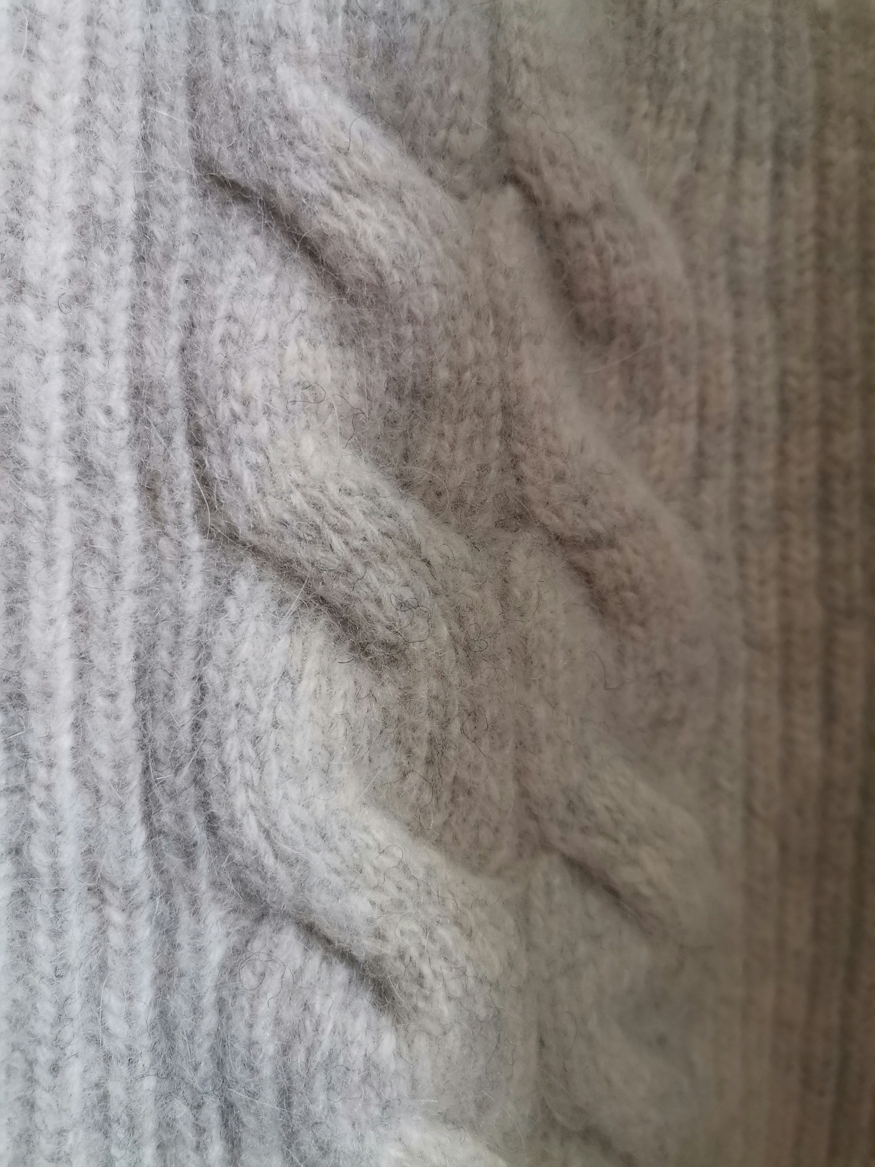 Close-up of a soft, light gray knitted fabric showcasing intricate cable patterns and ribbing. The texture highlights the craftsmanship of the material.