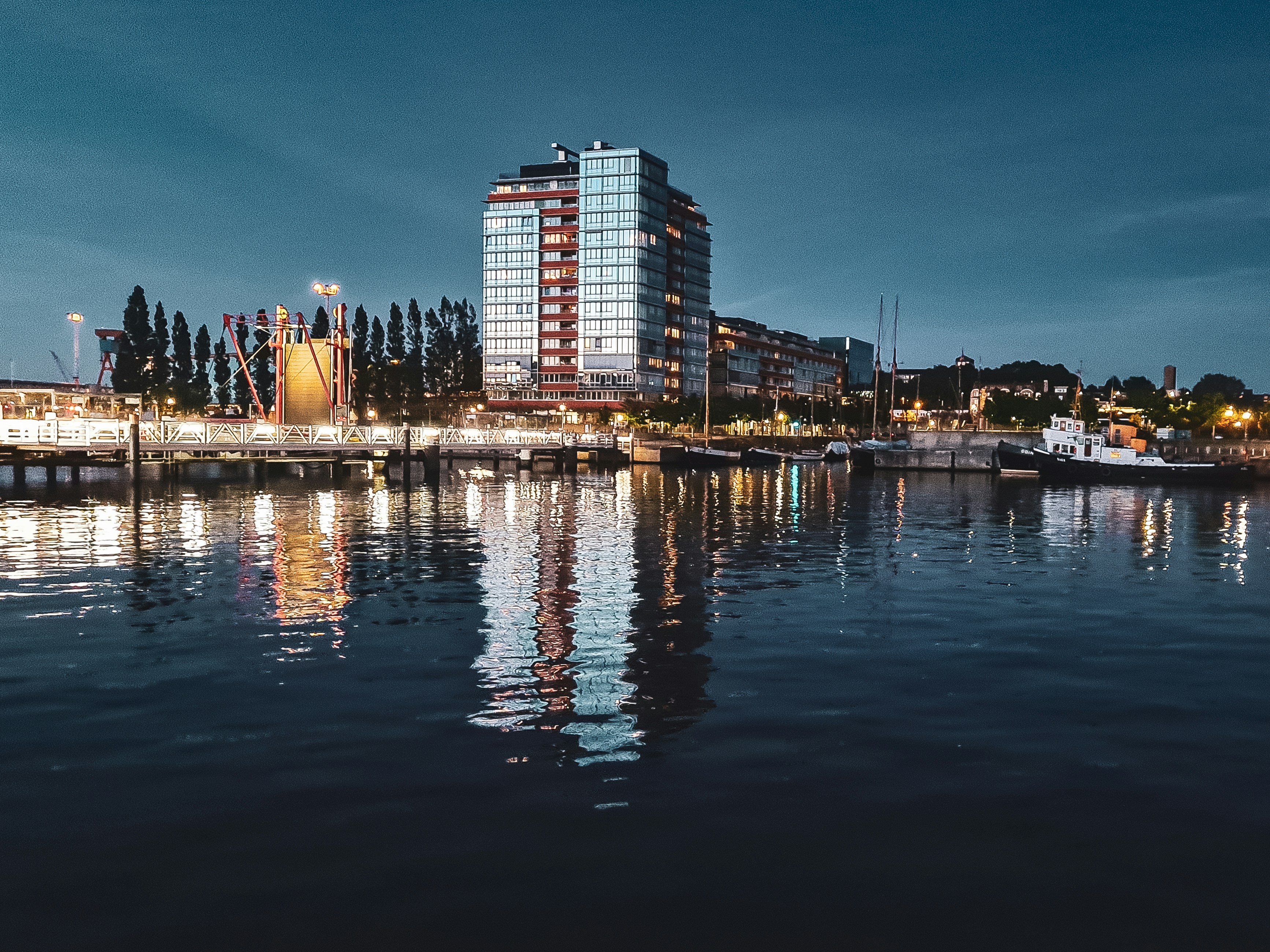 Modern waterfront building illuminated at dusk, reflecting on calm waters with boats docked nearby.
