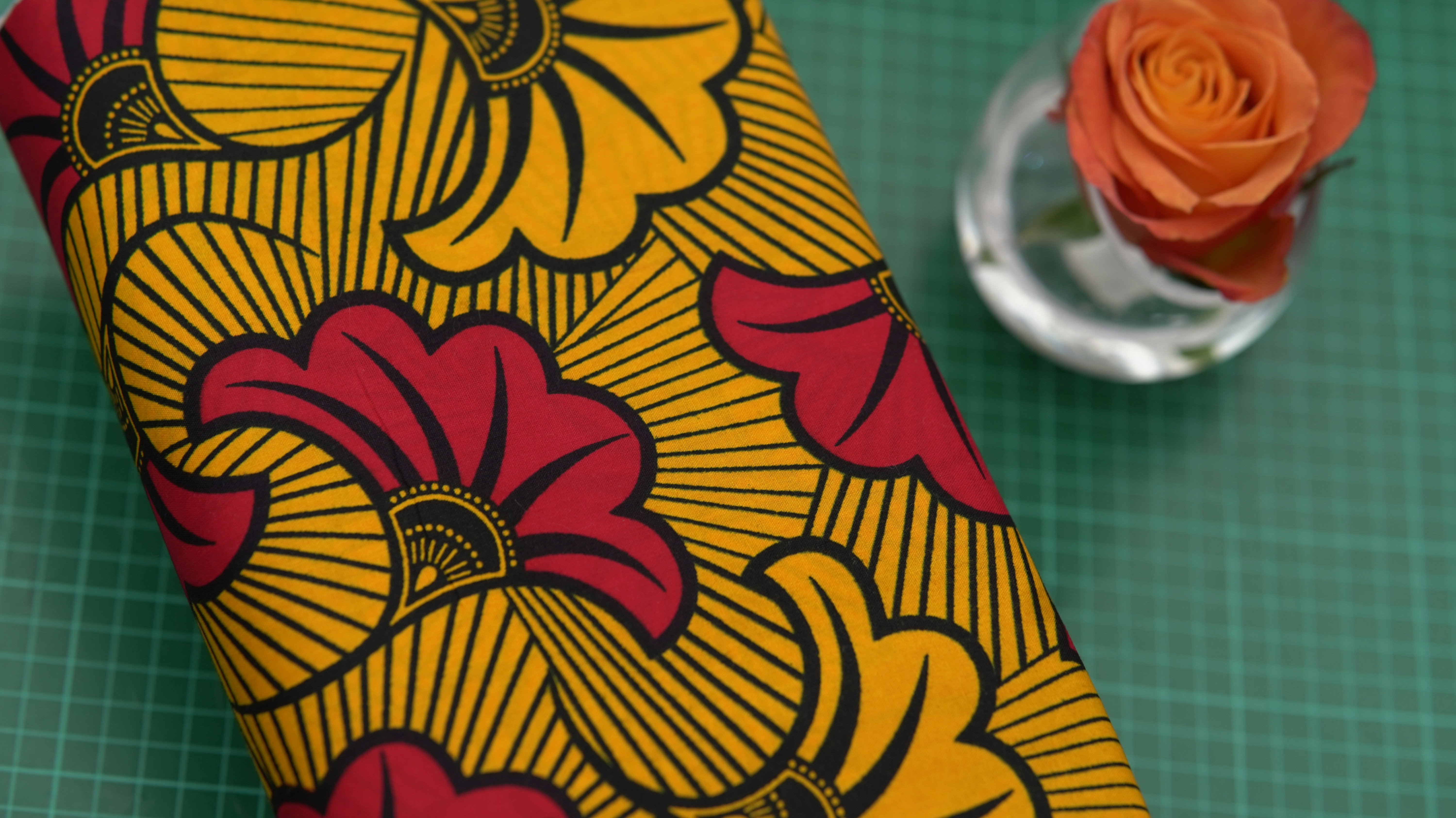 Colorful fabric with intricate floral patterns alongside a delicate rose in a glass vase. The composition highlights the blend of artistry and nature.